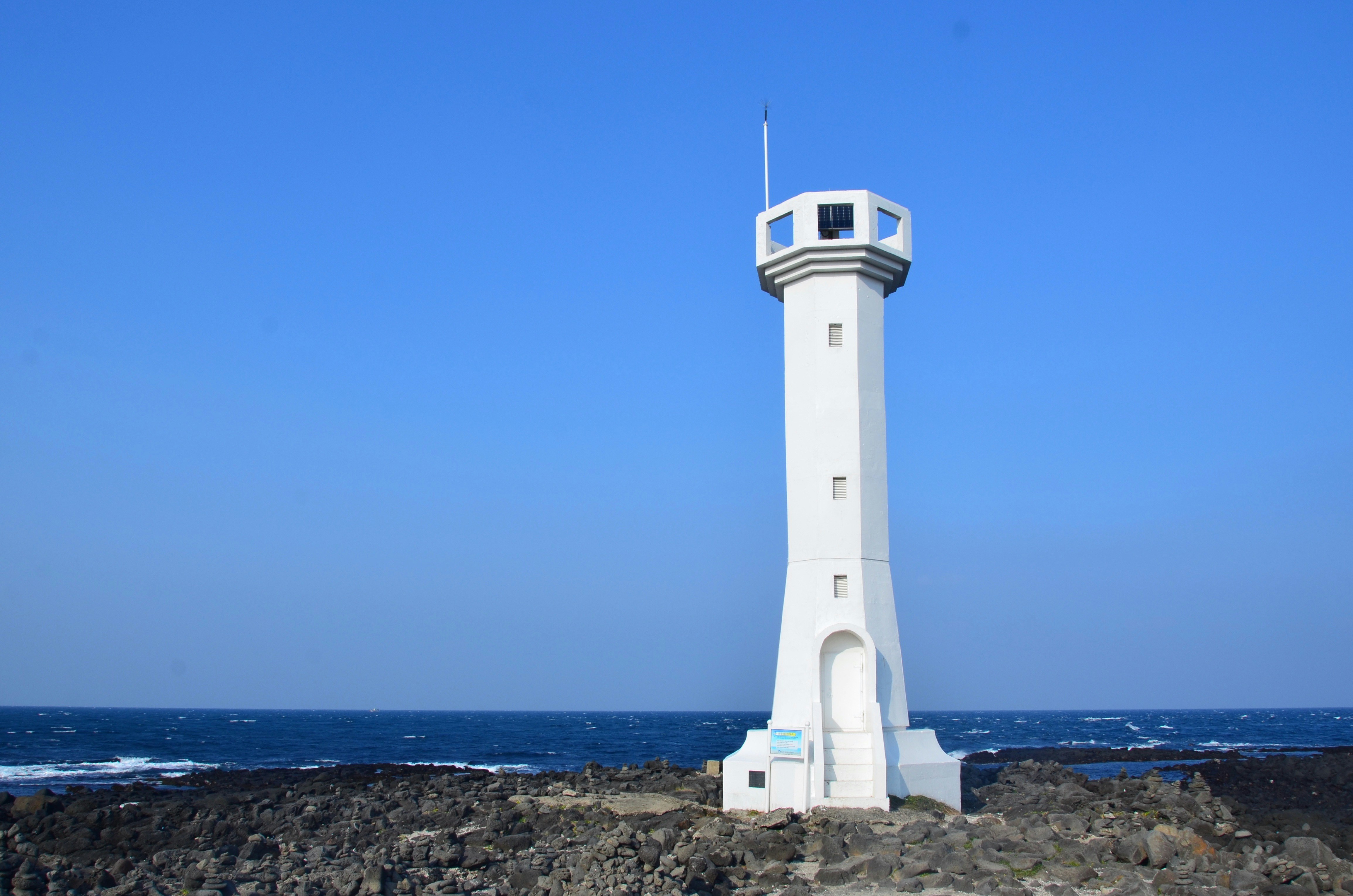 A white lighthouse stands tall on rugged rocks, contrasting against the deep blue sea and clear sky.