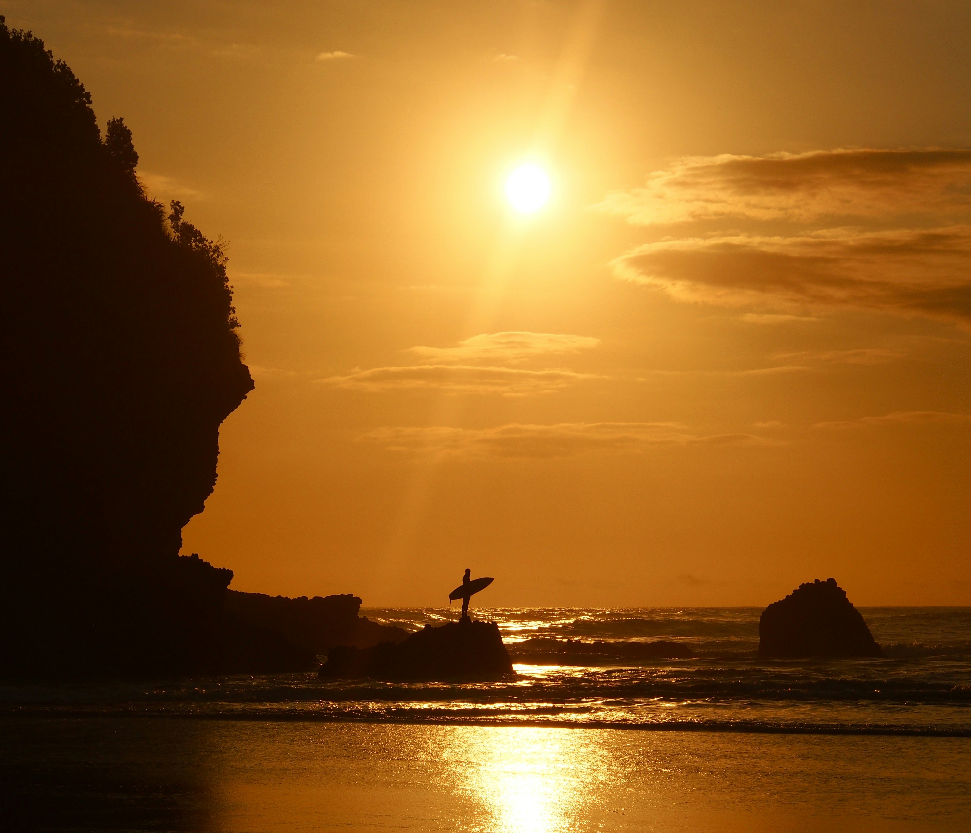 Silhouetted surfer poised on a rocky outcrop against a vibrant sunset, with golden hues reflecting on the water's surface.