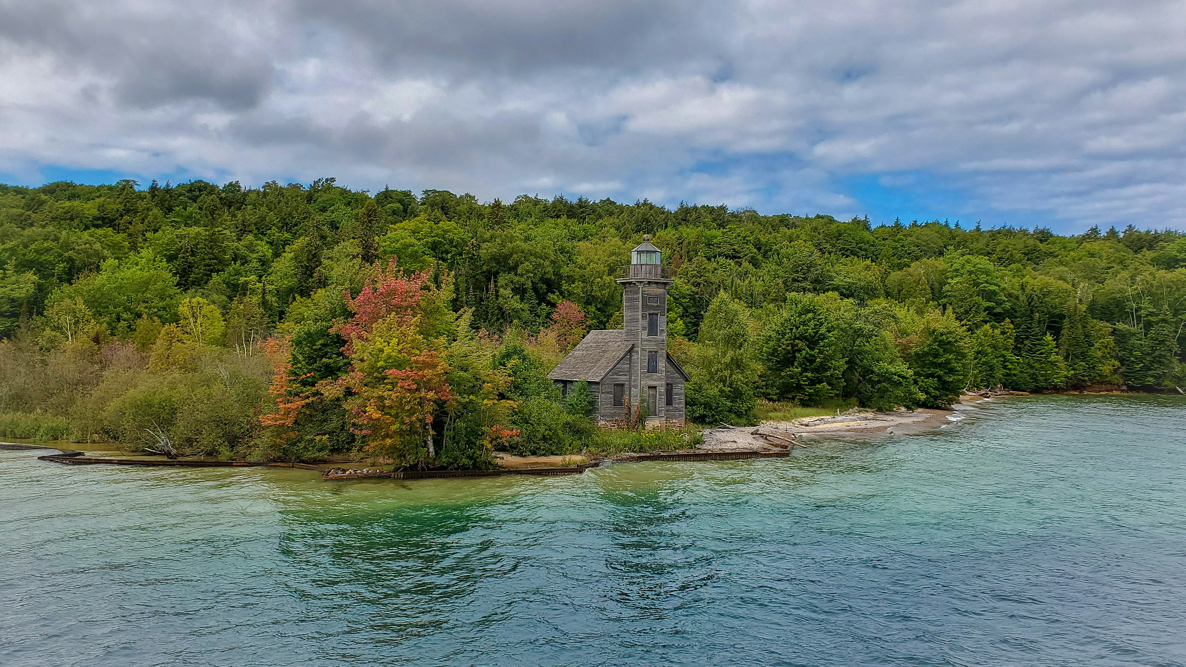 Abandoned lighthouse stands amidst vibrant autumn foliage on the shoreline, surrounded by calm waters and a lush forest backdrop.