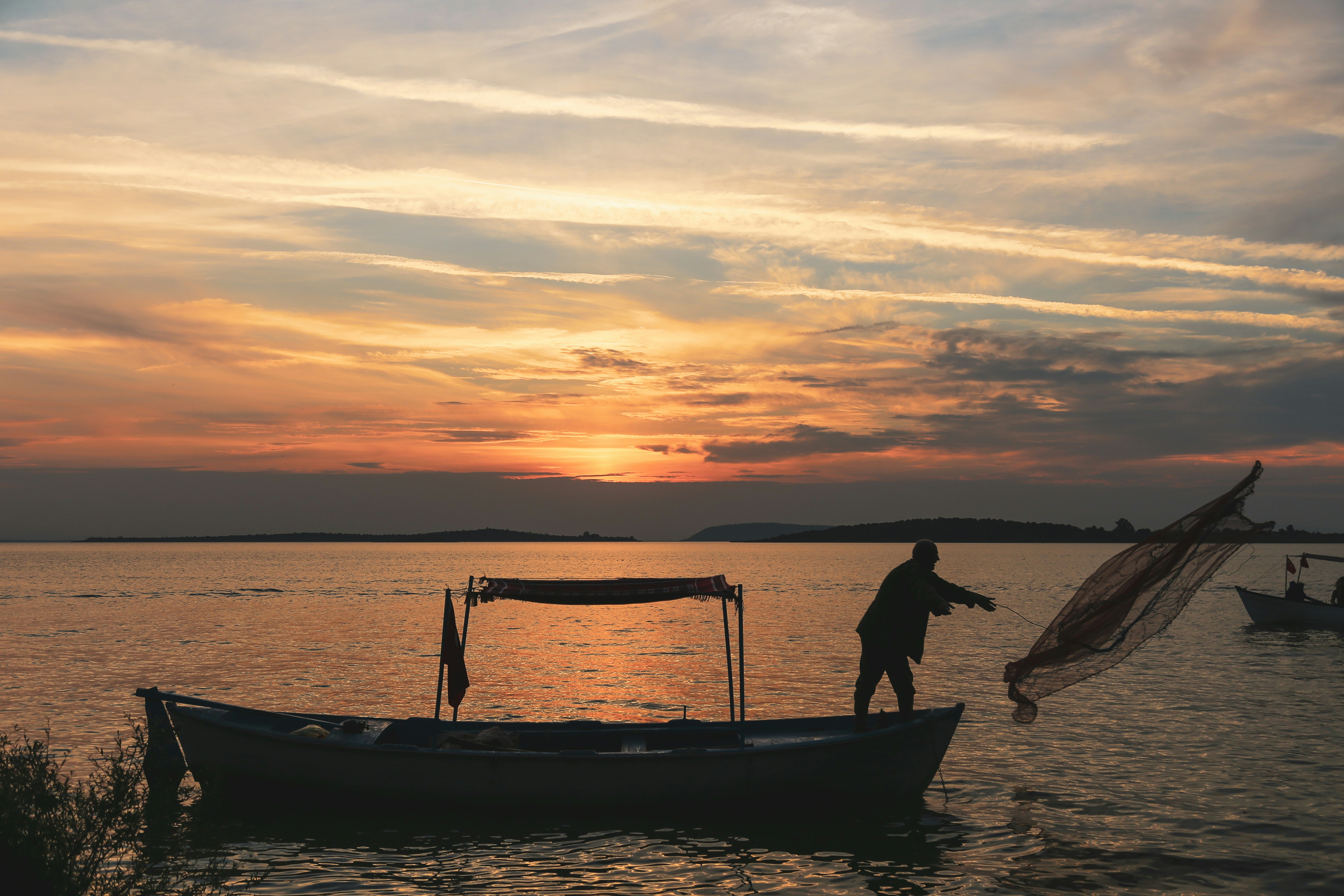Silhouette of a fisherman casting a net from a boat against a vibrant sunset sky over calm waters.