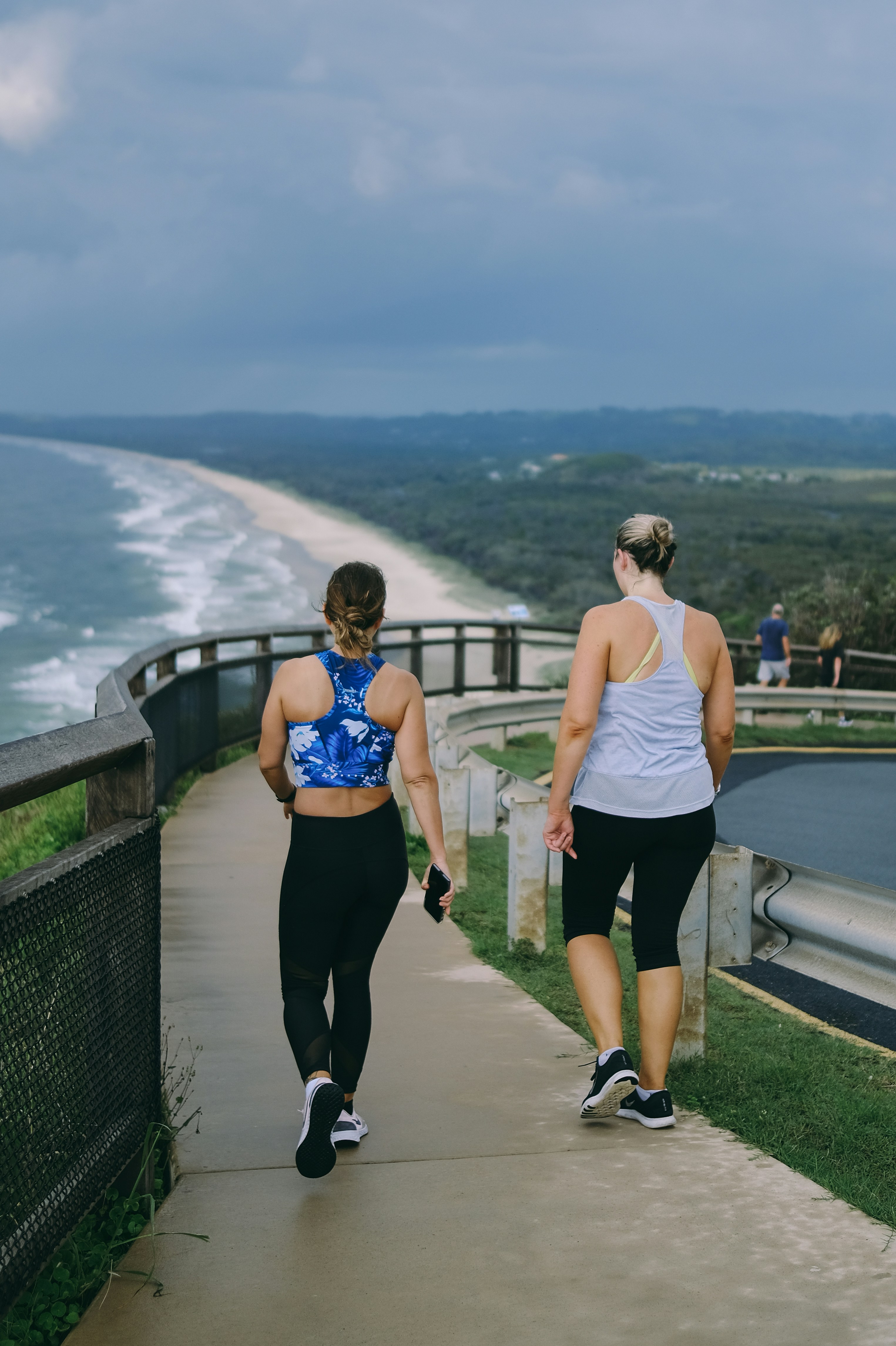 Two women walking along a coastal path, surrounded by lush greenery and ocean views. The scene captures a moment of connection with nature.