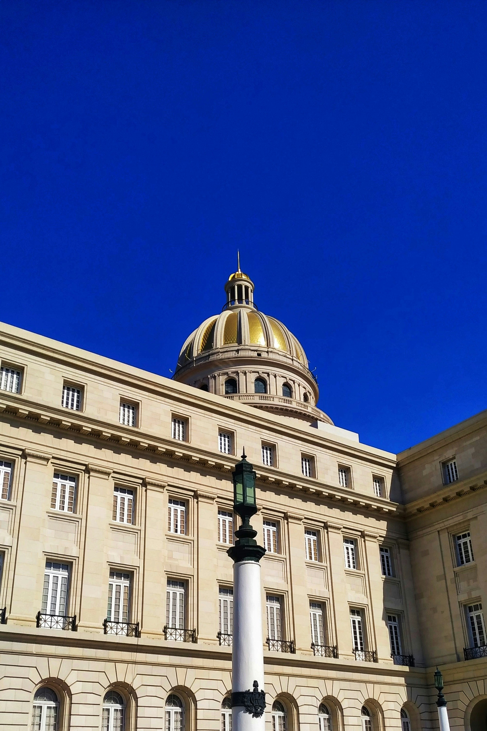 Neoclassical building with a gilded dome rises against a vivid blue sky, dominating the frame. A tall white column with a decorative lamp sits in the foreground, adding depth and architectural context.