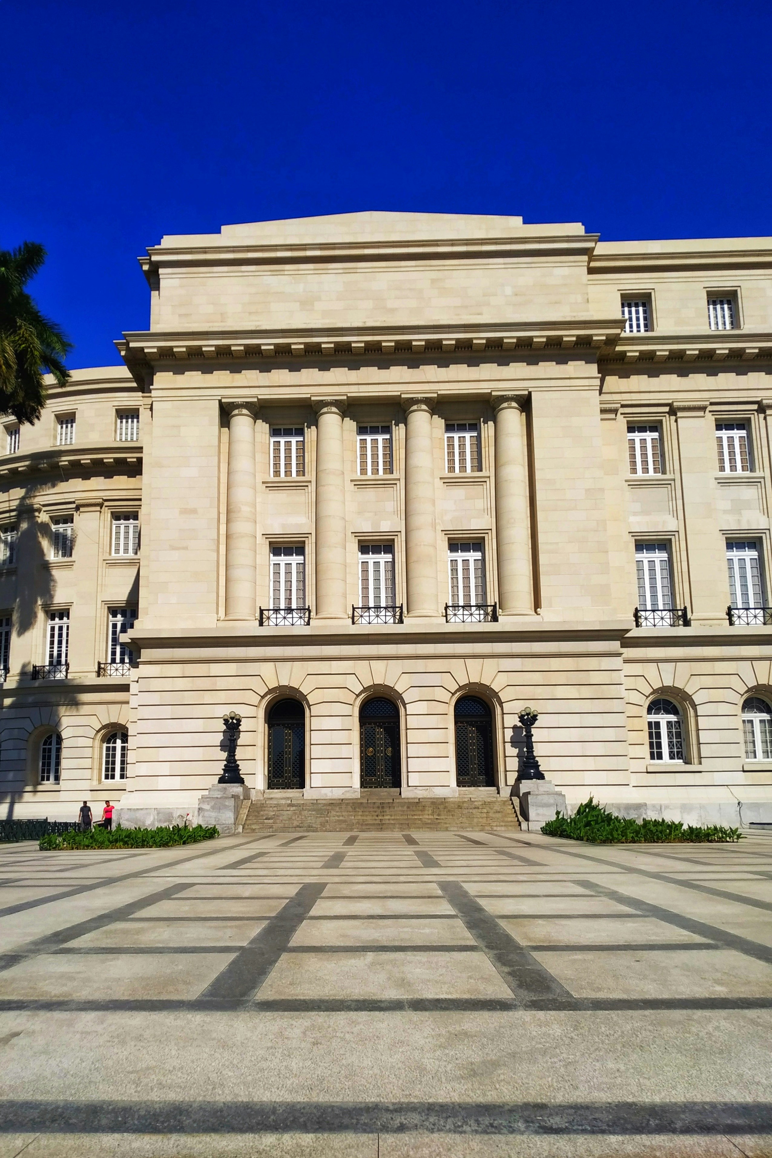 Imposing neoclassical building with grand columns and detailed stonework, framed by a clear blue sky. The entrance features ornate doors and statues flanking the steps.