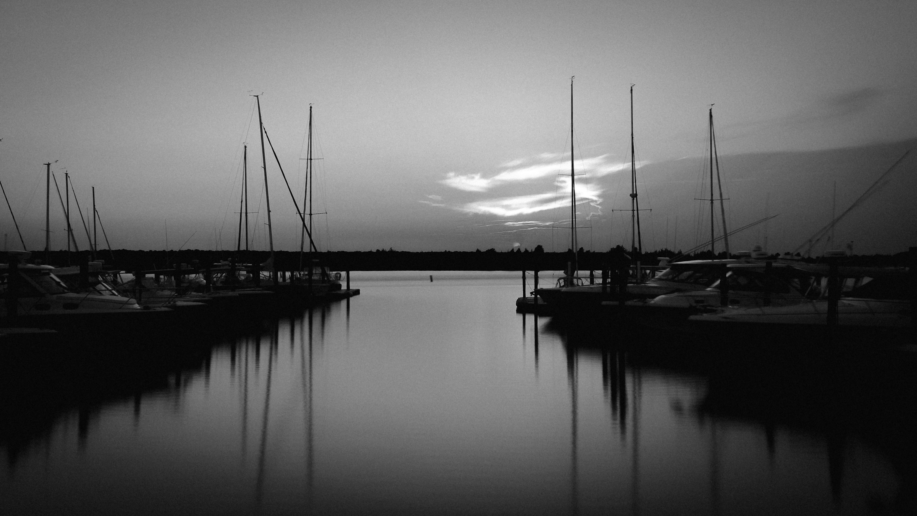 Black and white harbor scene featuring moored yachts silhouetted against a tranquil sunset. Reflections ripple across the still water.