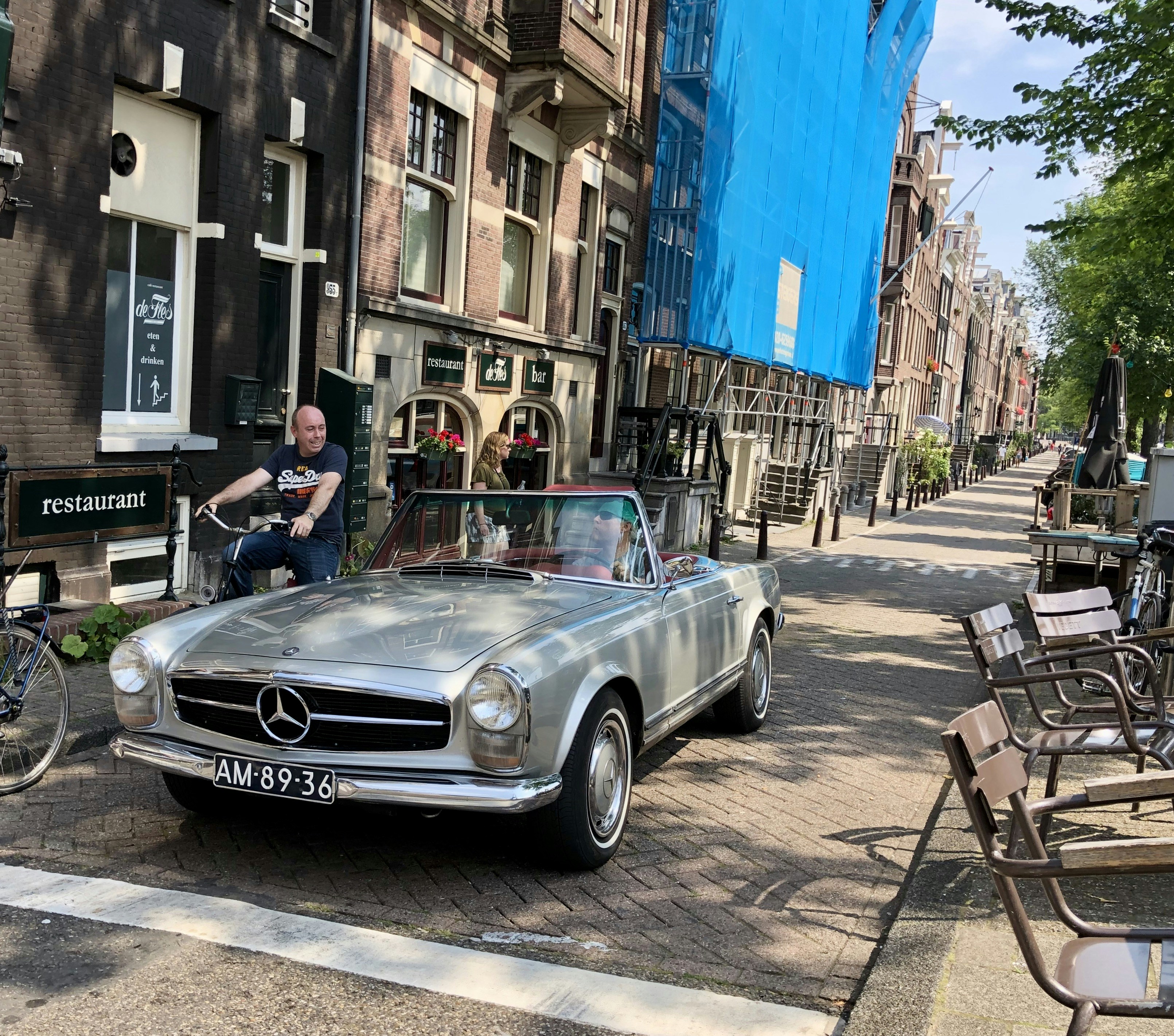 Vintage Mercedes convertible parked on a lively Amsterdam street, with bicycles and café seating nearby. Bright blue construction tarp adds a modern contrast.