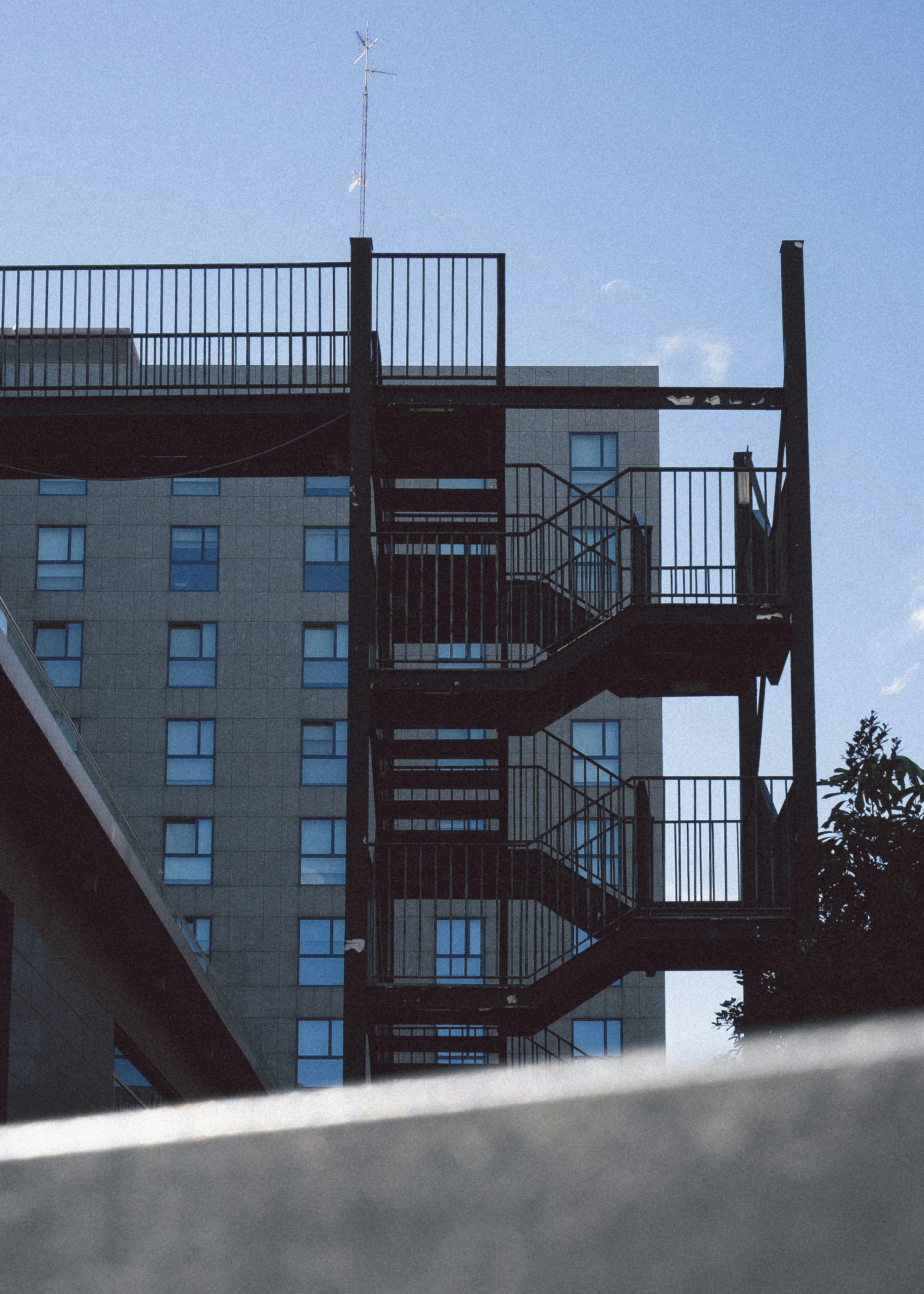 Metal fire escape spiraling up the side of a modern building, framed against a clear blue sky. The juxtaposition of industrial design and urban environment is striking.