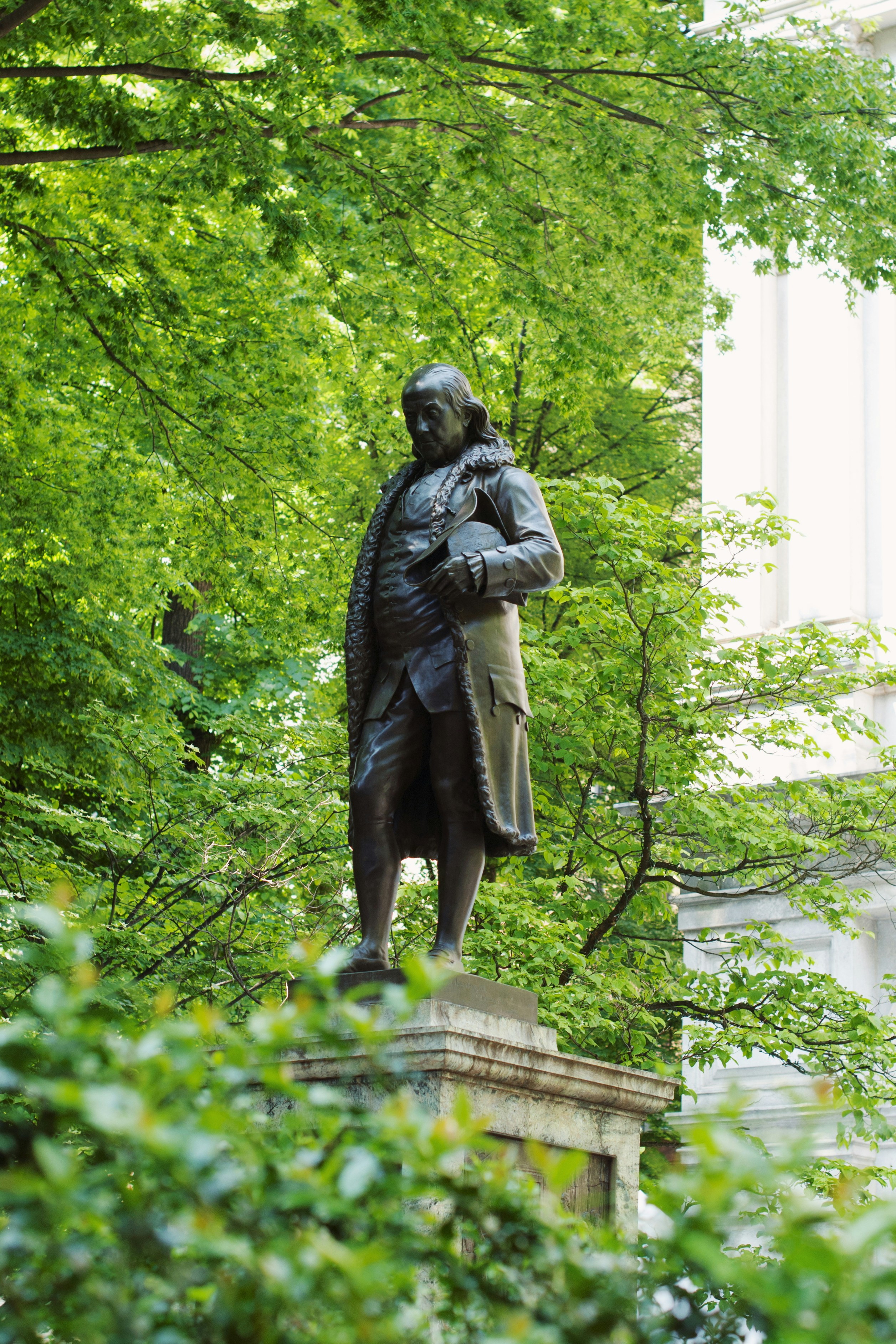 Bronze statue of a historical figure standing proudly on a pedestal, surrounded by lush green foliage in a serene park setting.
