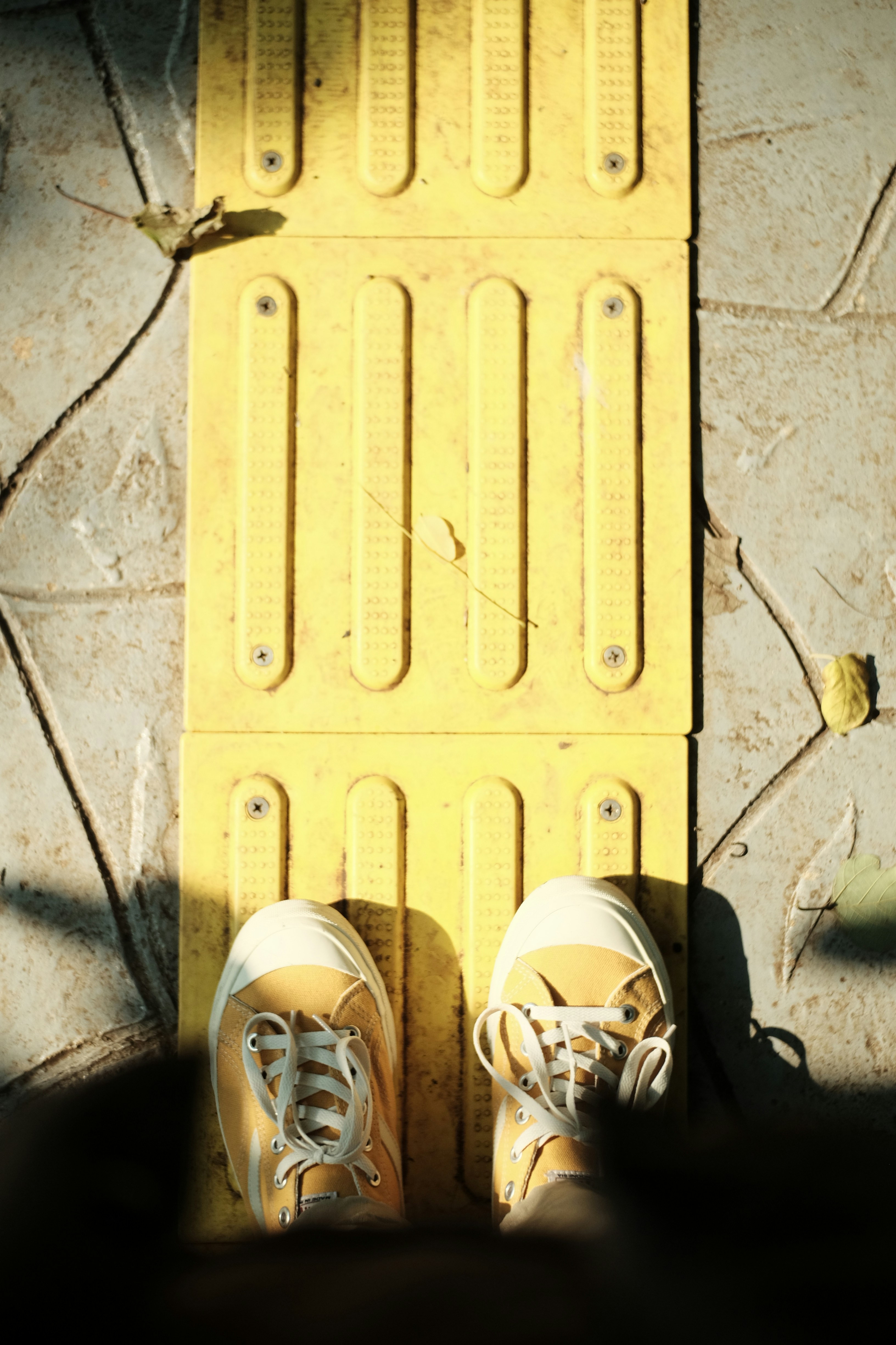 A pair of yellow sneakers viewed from above, standing on a bright yellow tactile paving strip along a sidewalk.