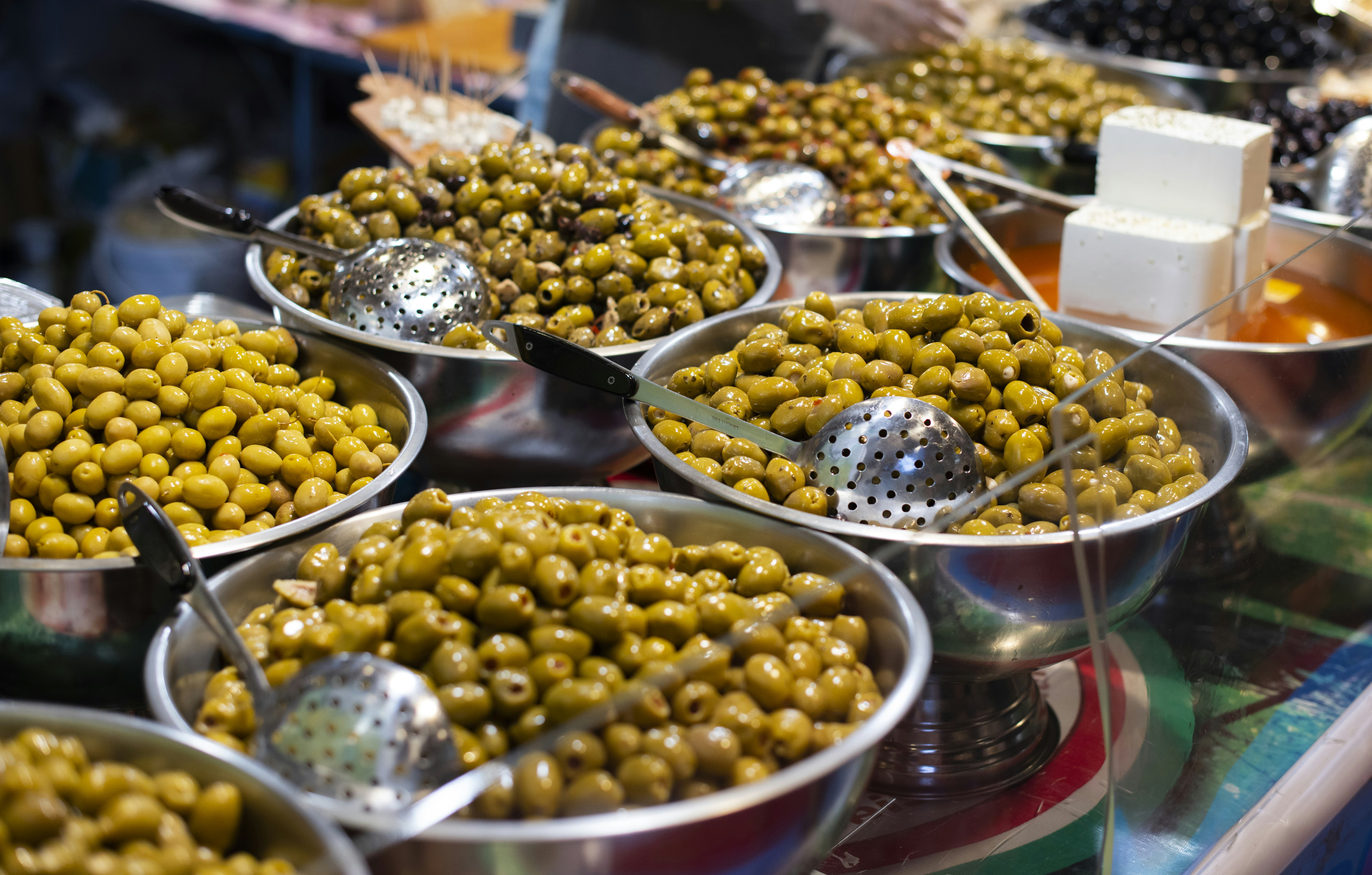 Bowls of green olives with stainless steel spoons displayed on a market stall.