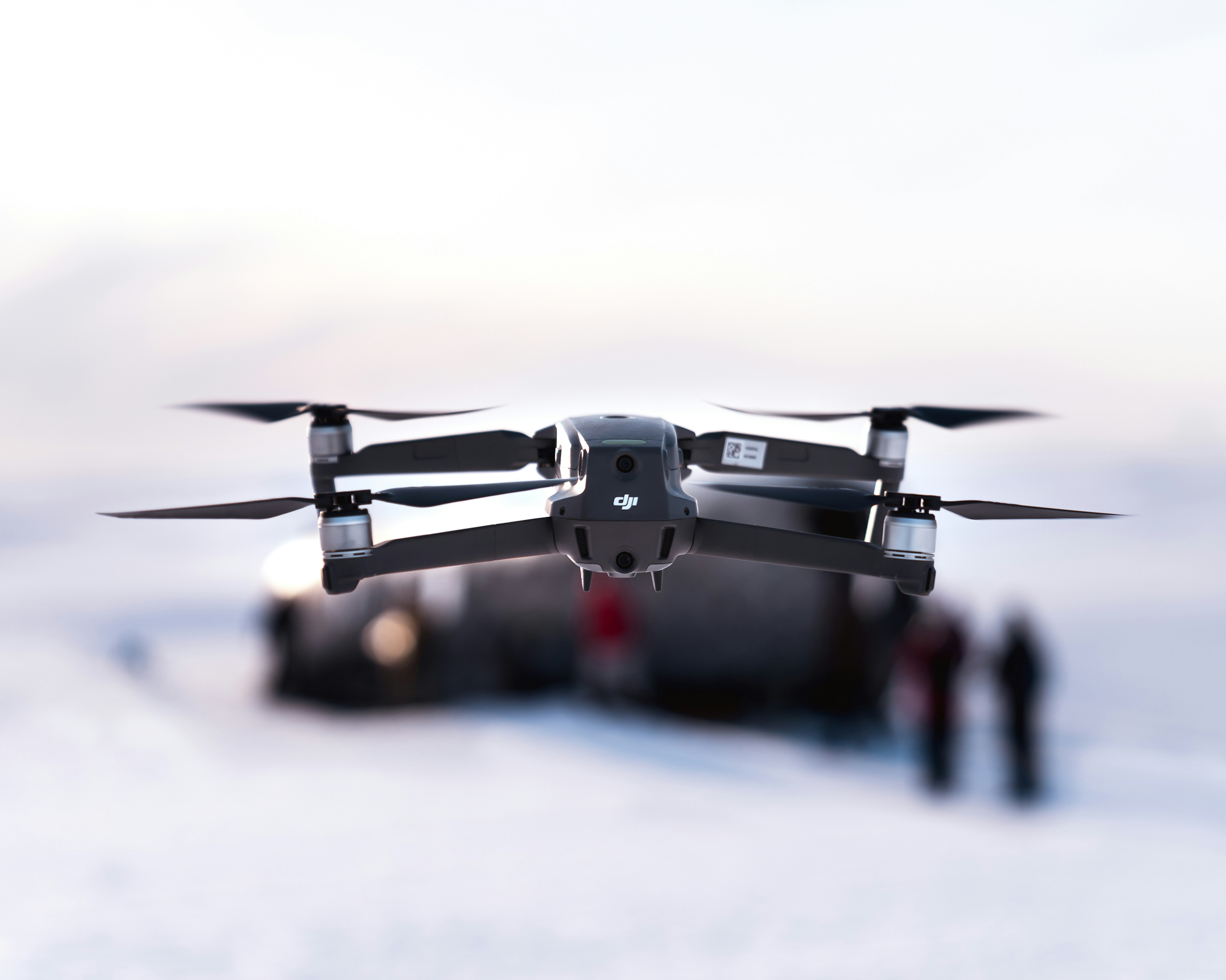 A sleek drone hovers in the foreground against a blurred backdrop of people and a structure in a snowy landscape. The scene captures the intersection of technology and nature.