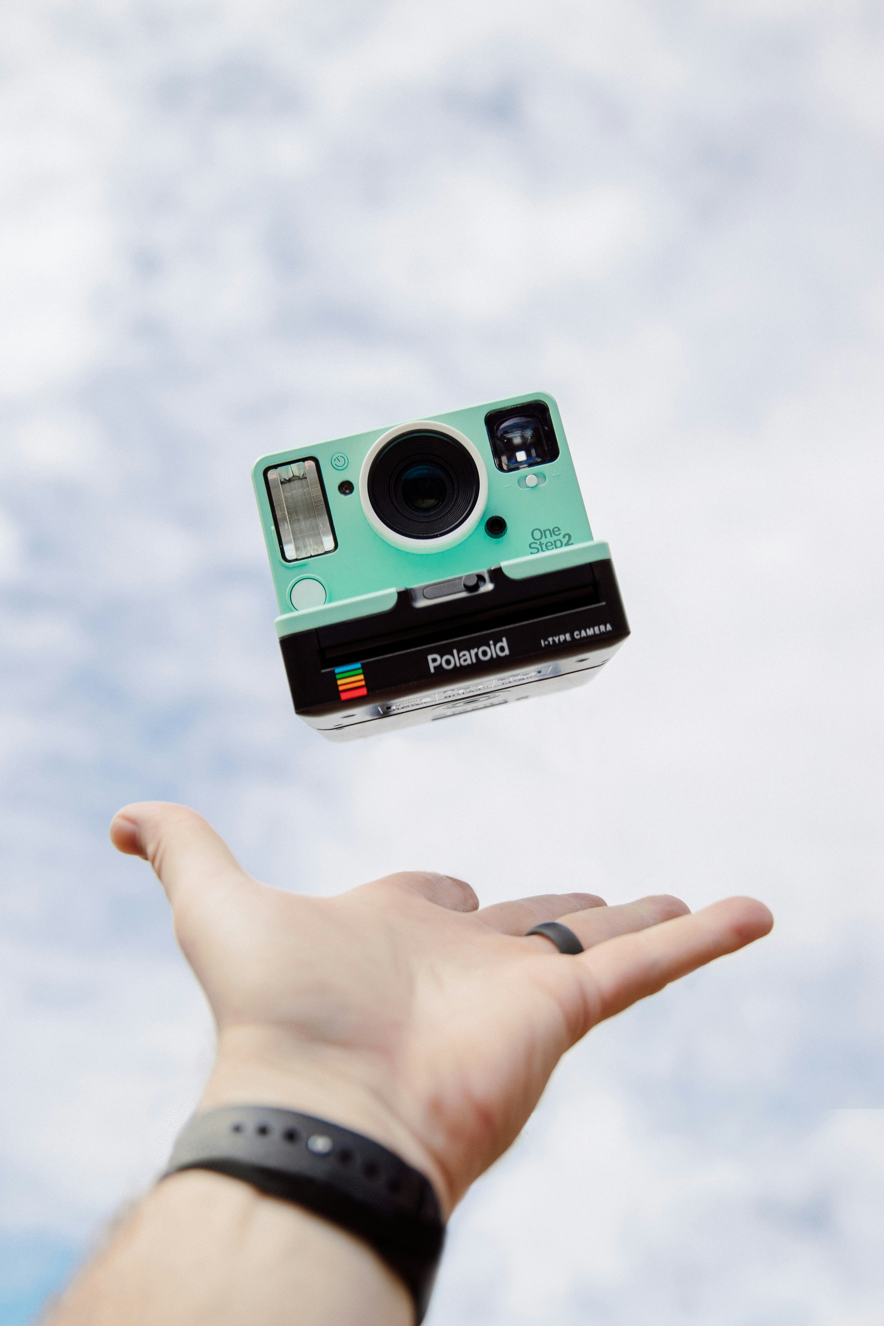 A Polaroid camera floats above an outstretched hand against a backdrop of soft clouds. The scene captures the essence of retro photography.