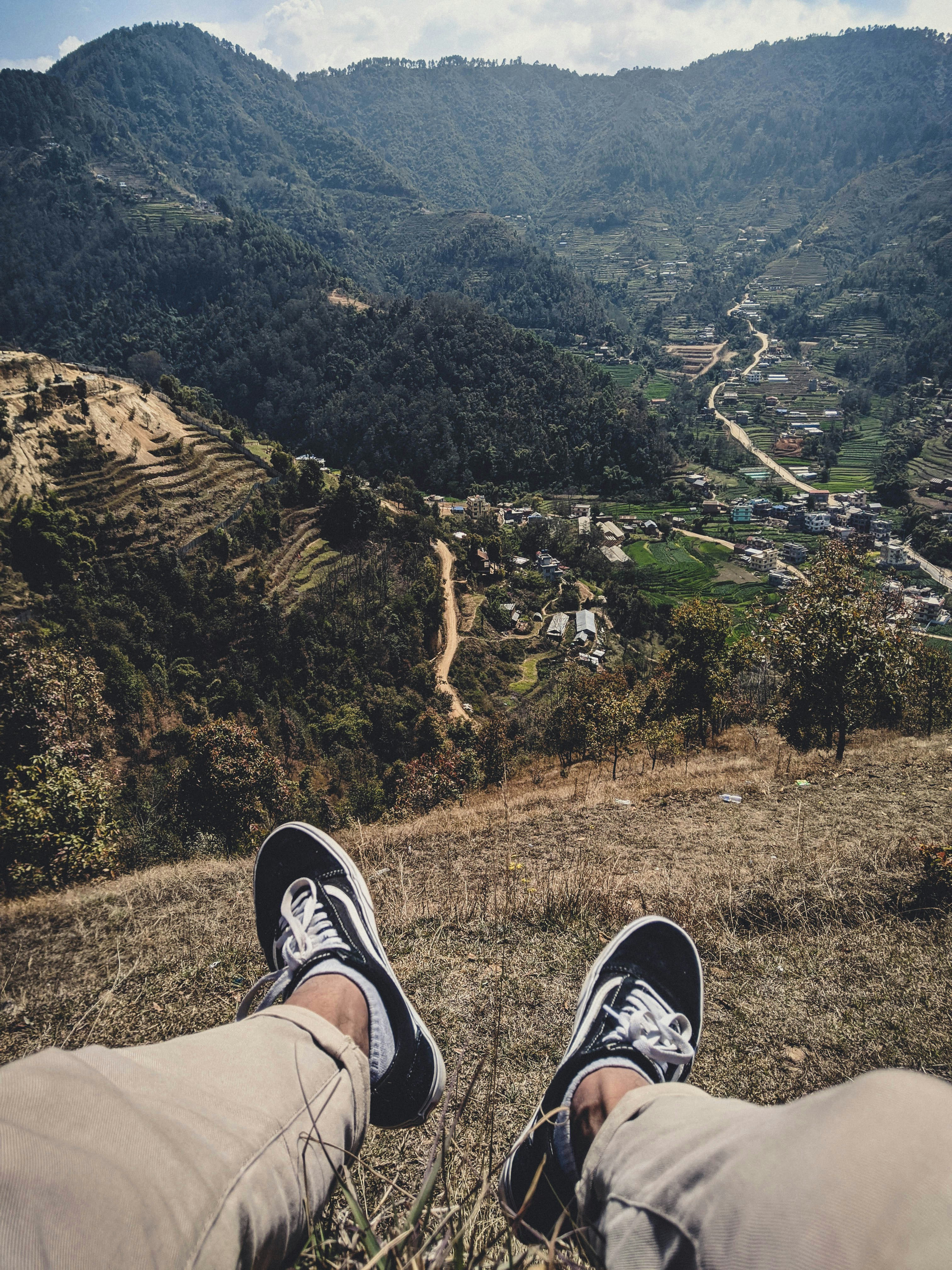A pair of sneakers resting on a grassy slope, overlooking a lush valley with terraced fields and a winding road below.