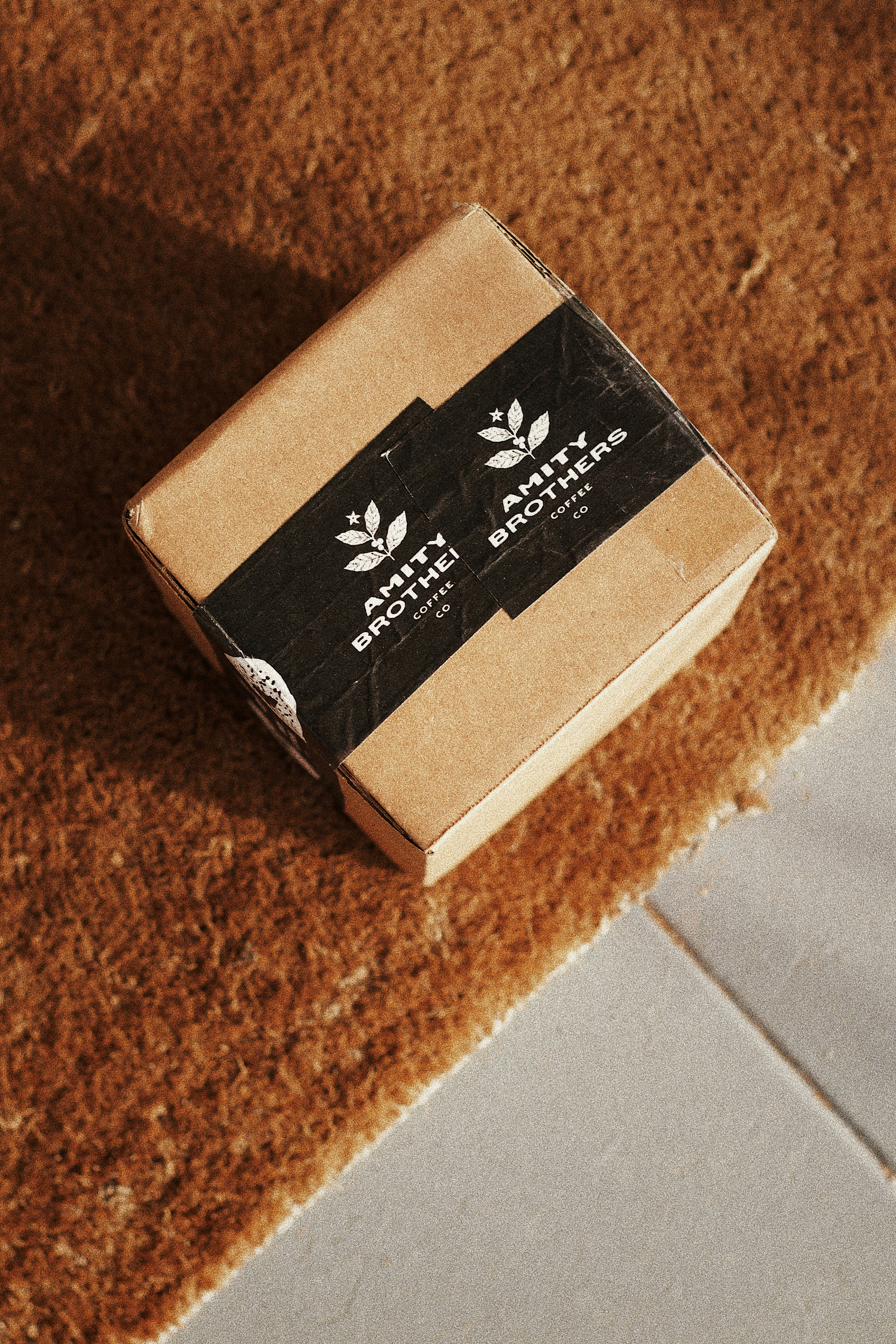 Cardboard parcel sealed with black logo tape rests on a rust-orange textured rug.