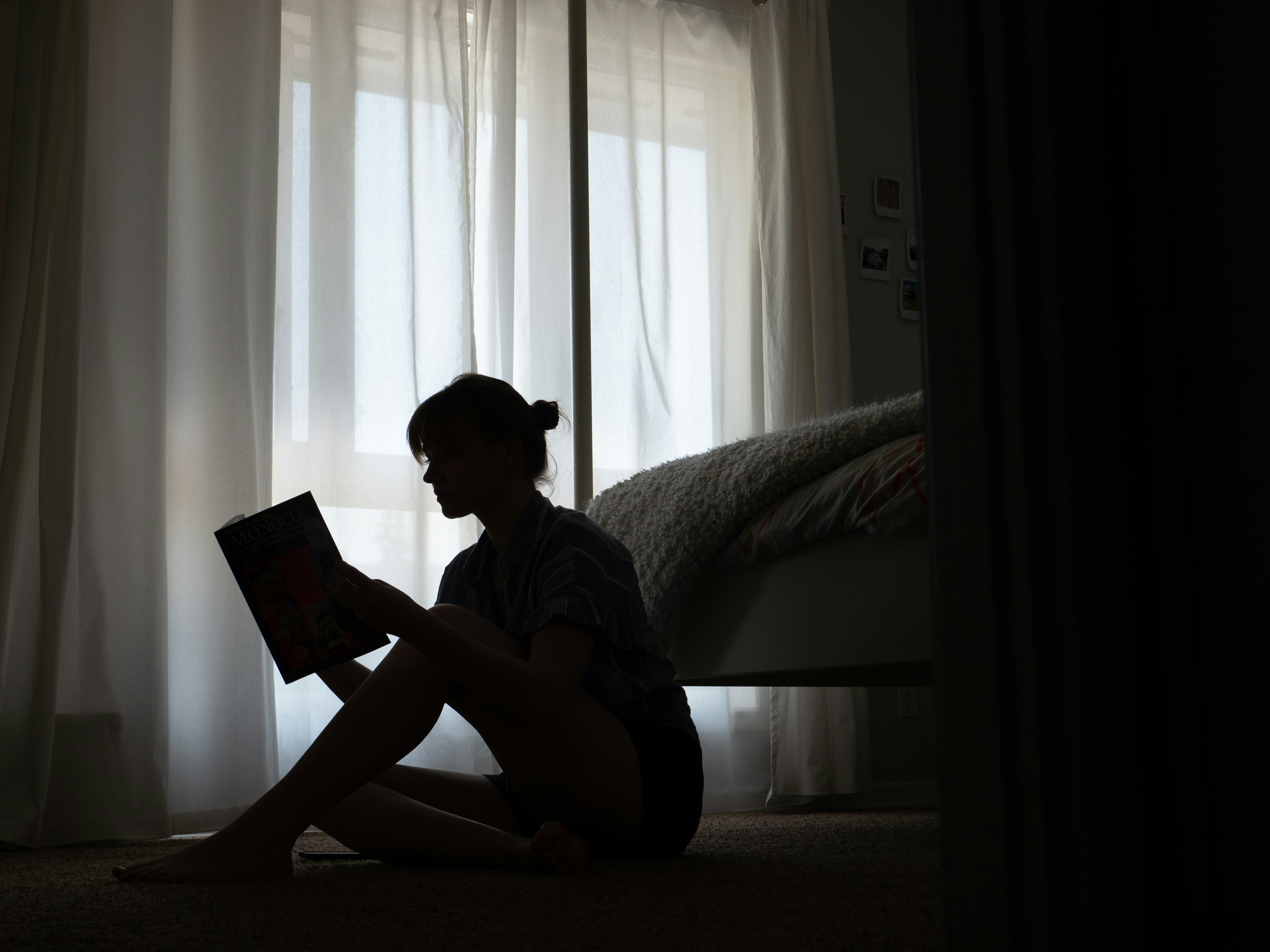 Silhouette of a person reading a book in a softly lit room with sheer curtains.