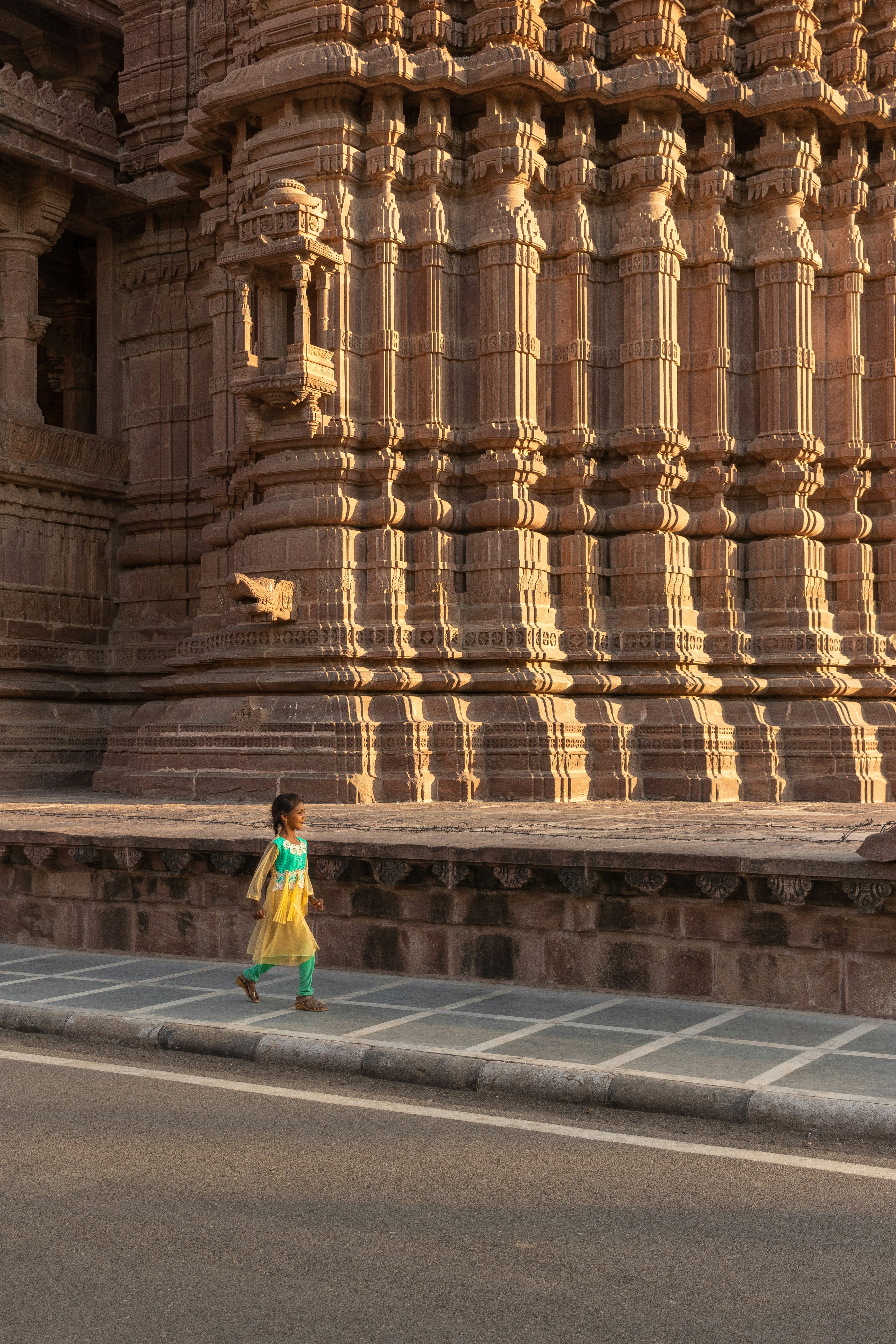A young girl walks along a stone pathway next to intricately carved temple walls, illuminated by warm sunlight.
