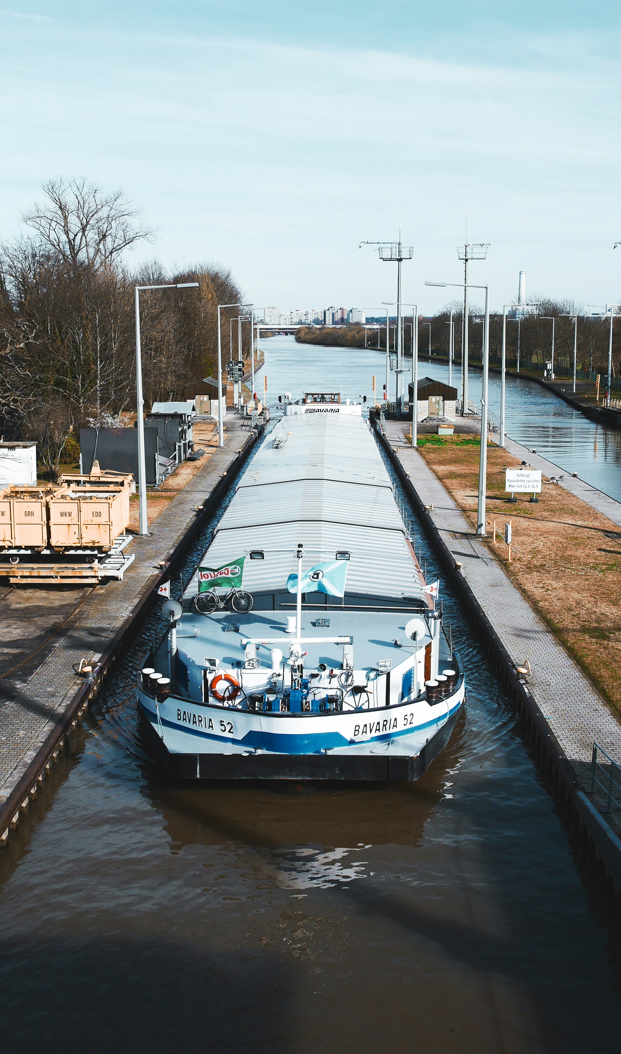 Cargo vessel Bavaria 52 gliding through a narrow canal lined with trees and utility poles.