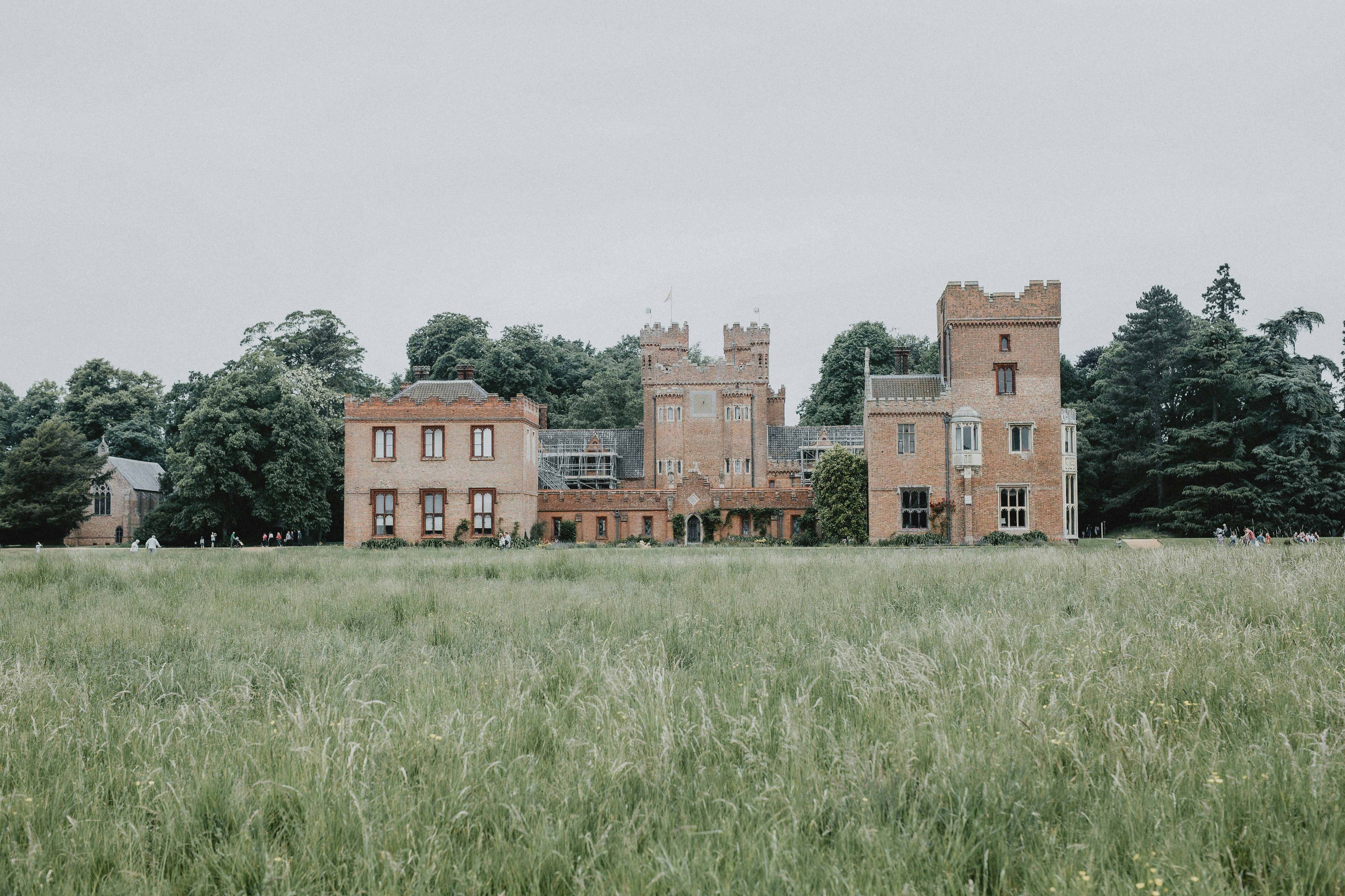 Historic brown brick building surrounded by lush green grass and towering trees under an overcast sky.