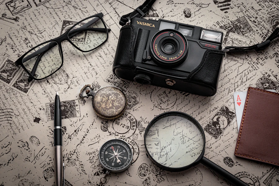 A Yashica film camera surrounded by eyeglasses, a compass, a magnifying glass, a pen, a pocket watch, and a brown wallet on a map-patterned surface. The arrangement has a nostalgic travel-and-exploration feel.