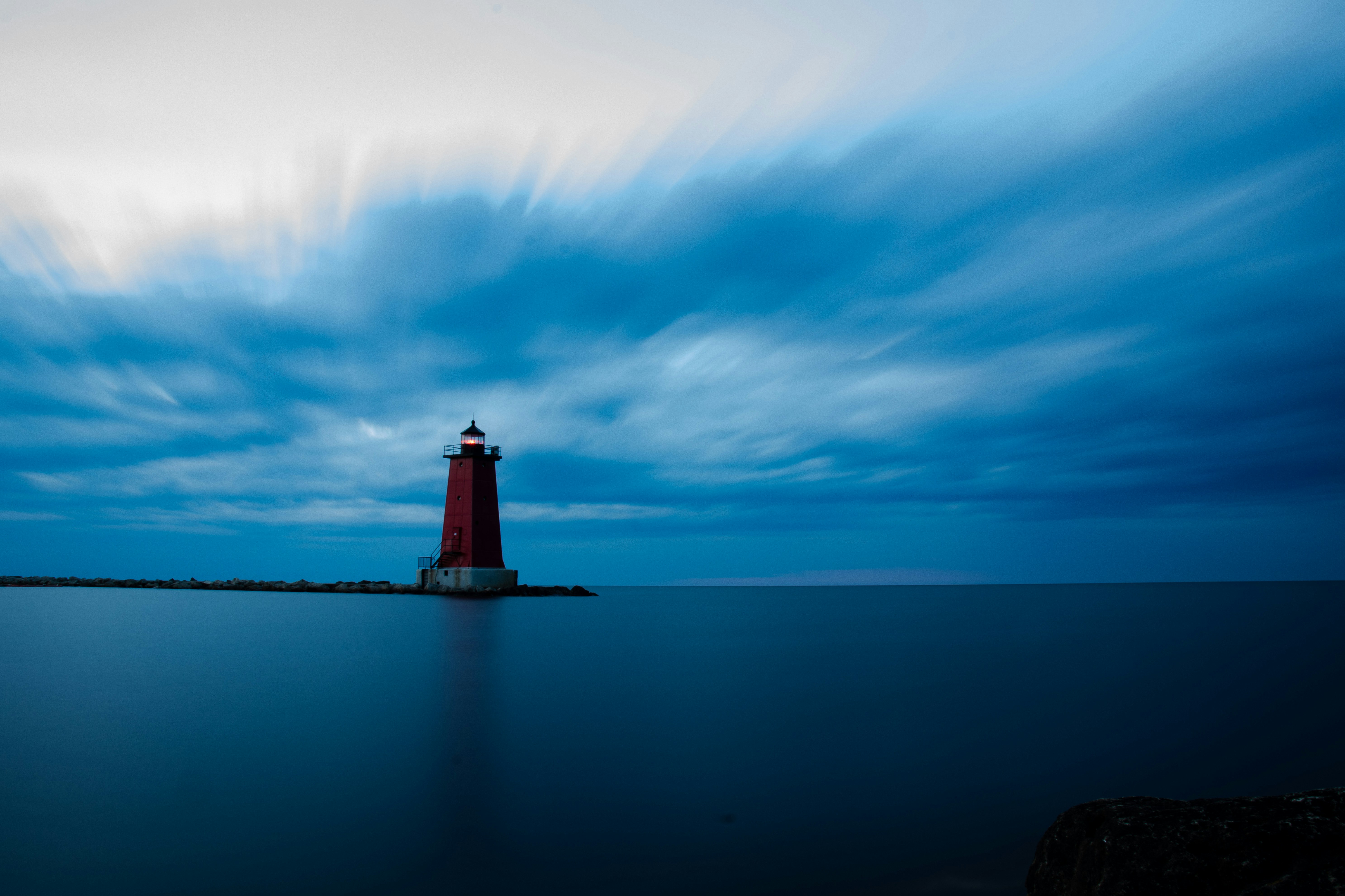A vibrant red lighthouse stands resilient against a moody sky, surrounded by calm waters at dusk.