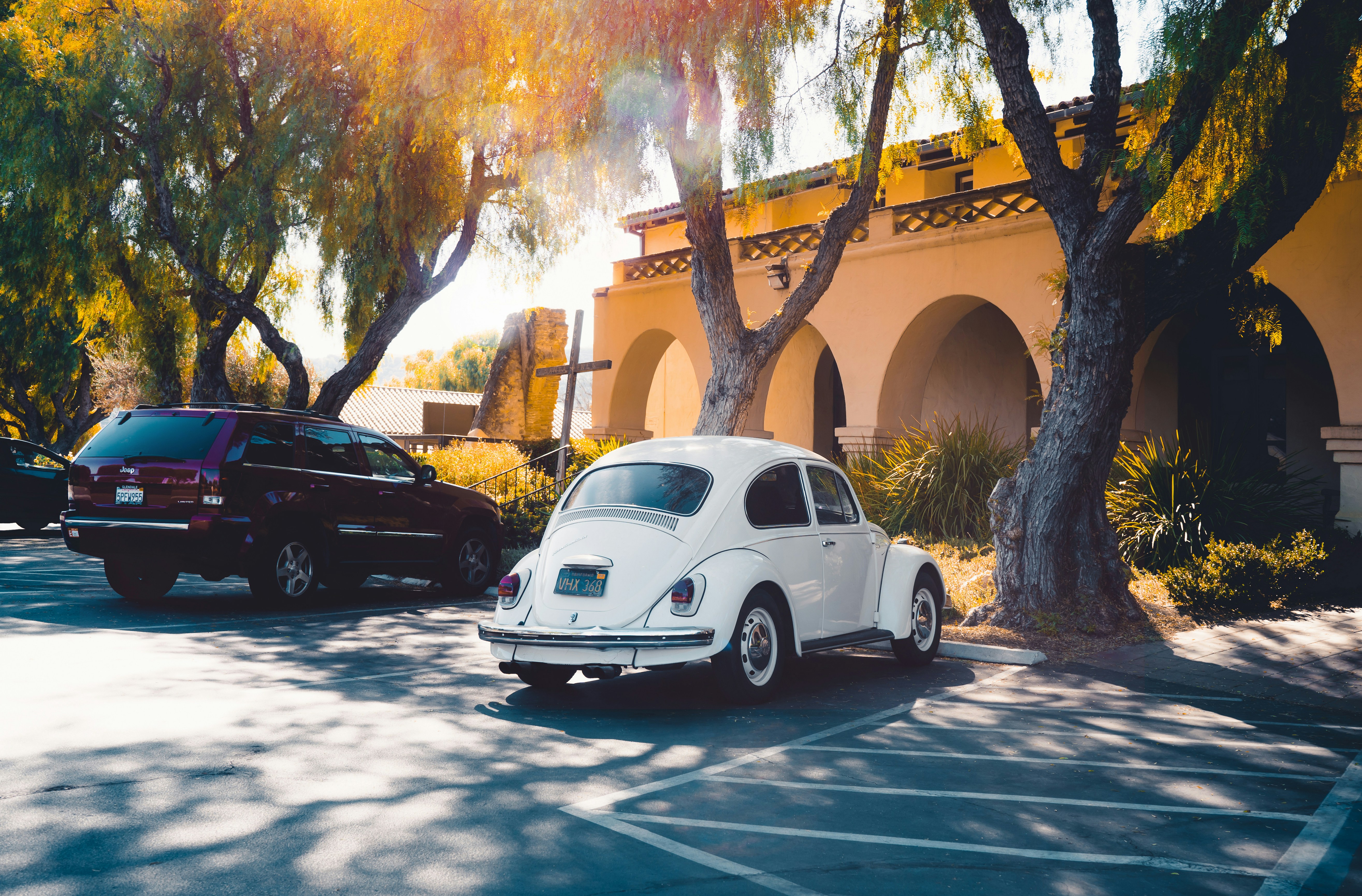 Vintage white Volkswagen Beetle parked under sunlit trees in a serene lot, showcasing a blend of nostalgia and contemporary architecture.