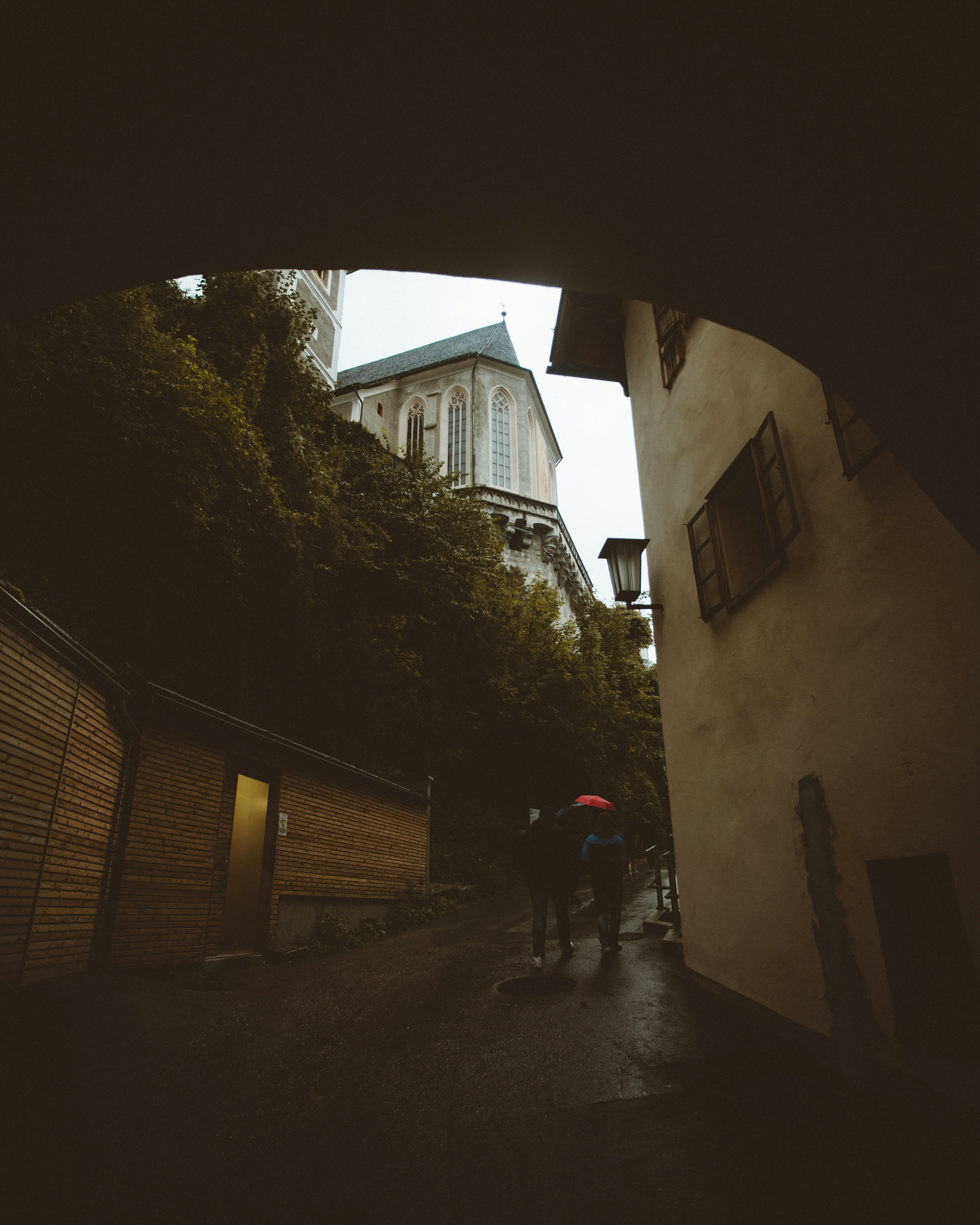 Two figures walking under an archway on a rainy day, with a historic church visible in the background amidst lush greenery.