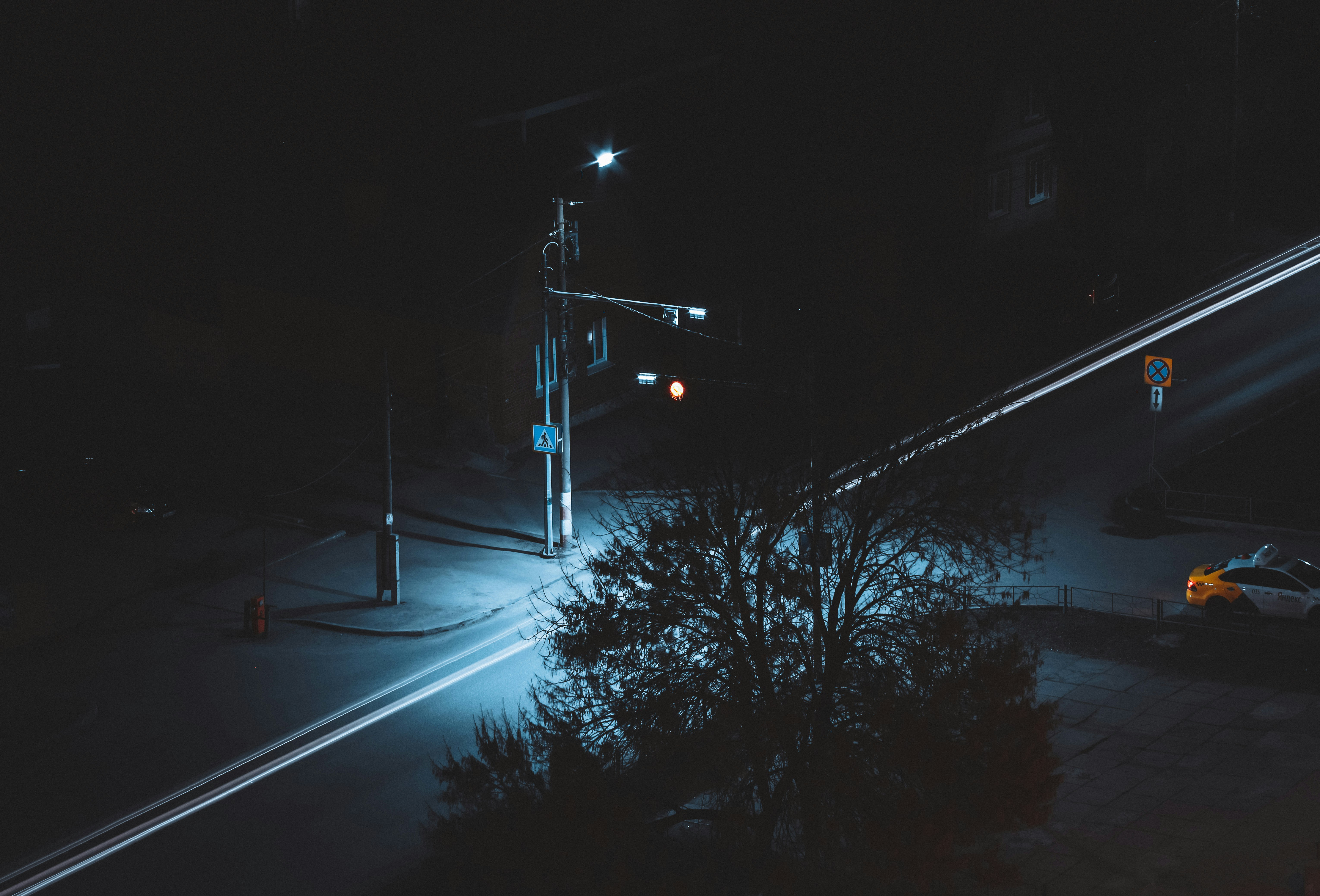 A dimly lit urban intersection illuminated by streetlights, with a solitary tree casting shadows on the pavement. A taxi is visible in the background, adding a touch of movement to the stillness.