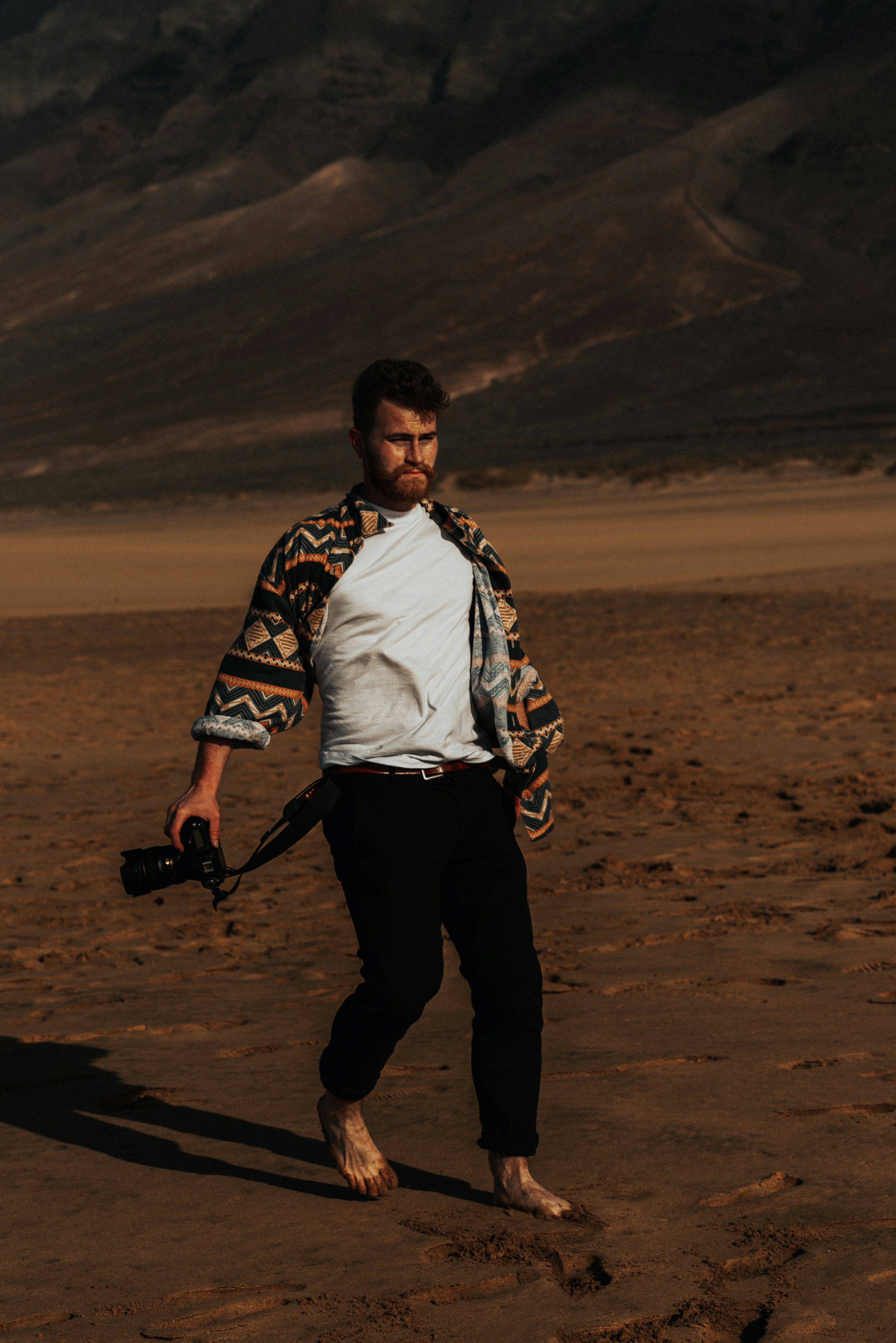 A man walks barefoot across a sandy desert landscape, holding a camera, dressed in a patterned jacket and plain shirt. The backdrop features distant mountains under a warm, golden light.