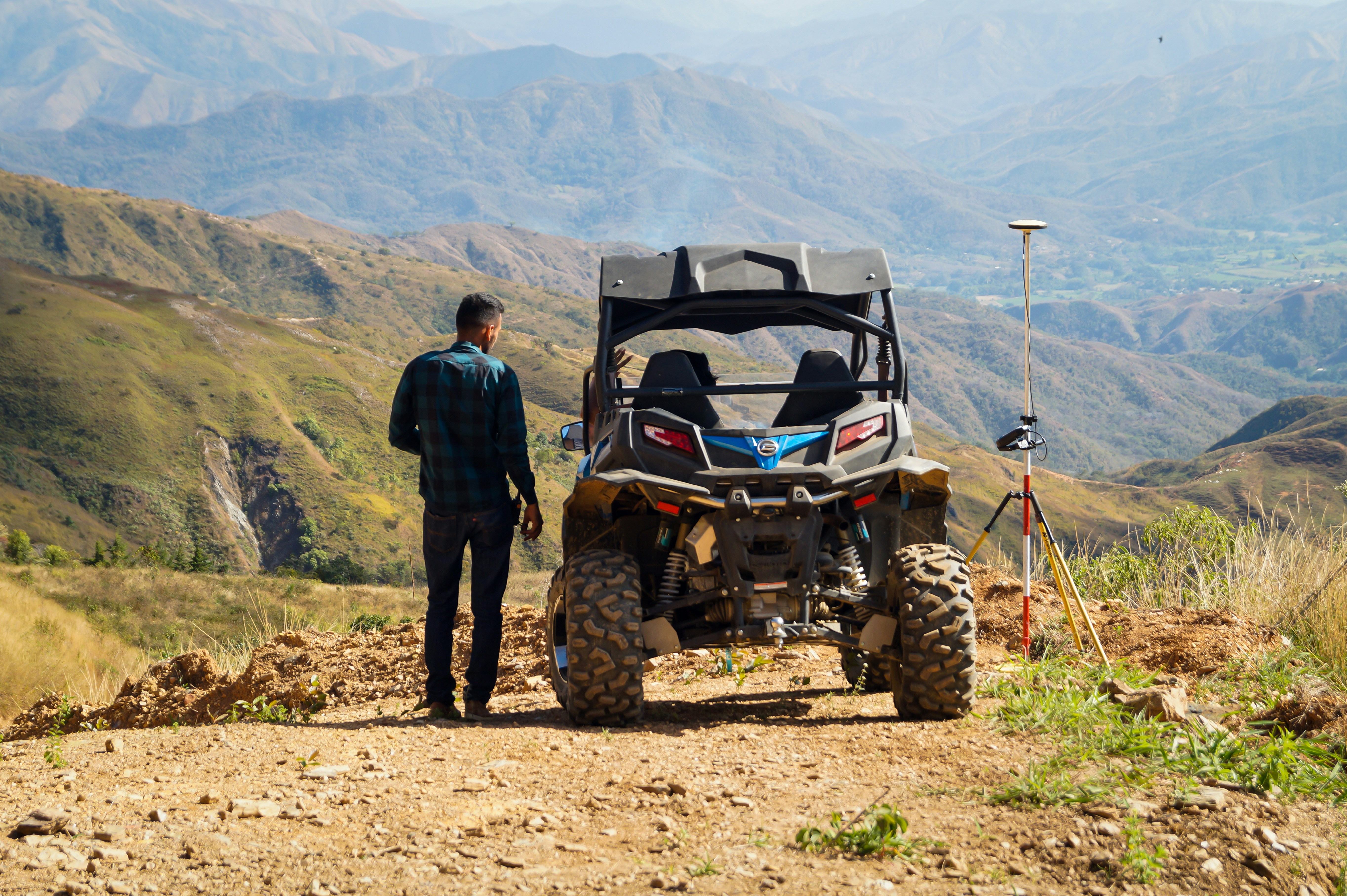 A person stands beside an all-terrain vehicle on a rugged mountain path, gazing into the expansive landscape. A surveying instrument is positioned nearby.