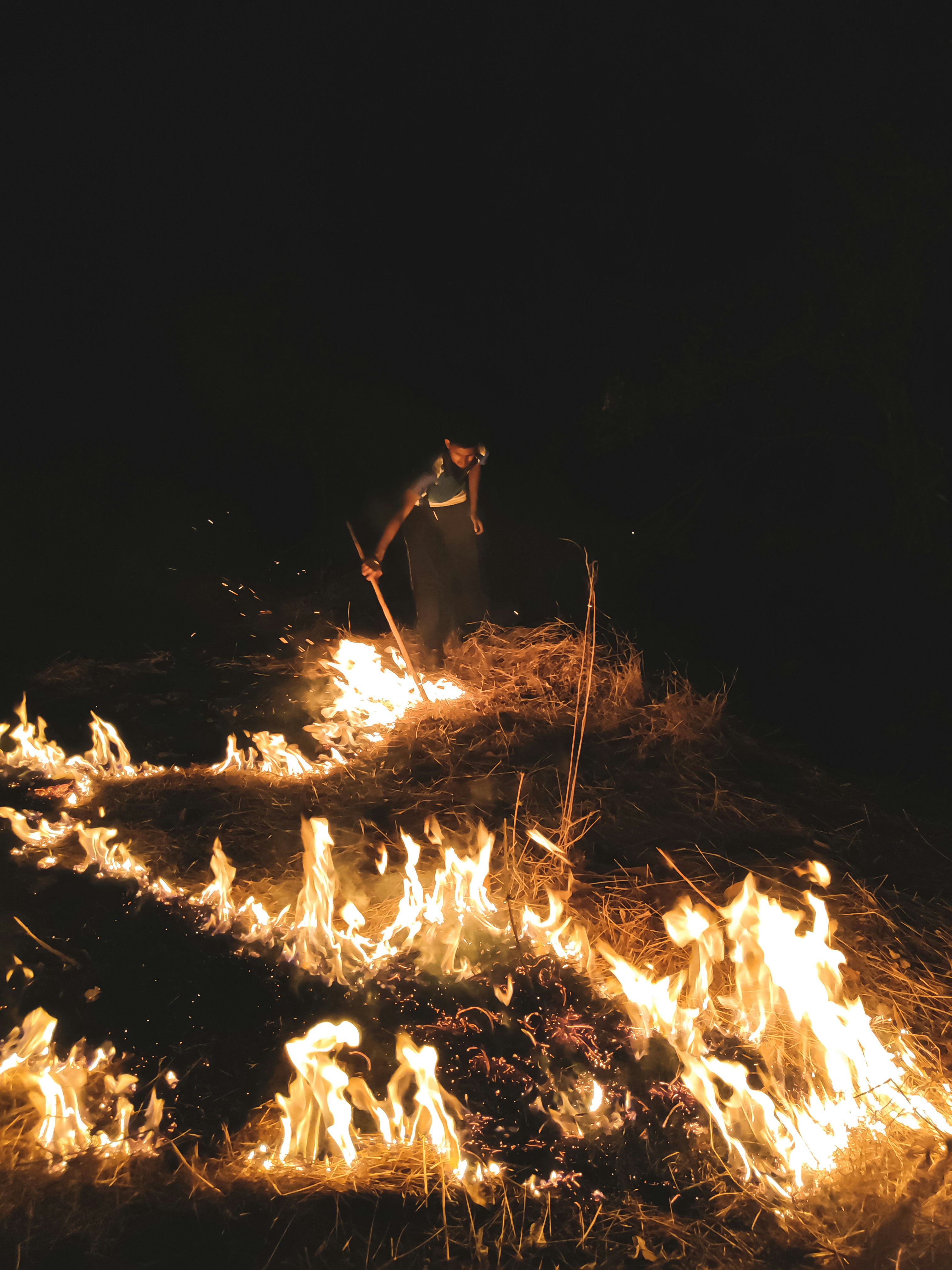 Night photograph of a lone figure tending a line of flames across a dry field. Sparks rise as the fire lights the scene.
