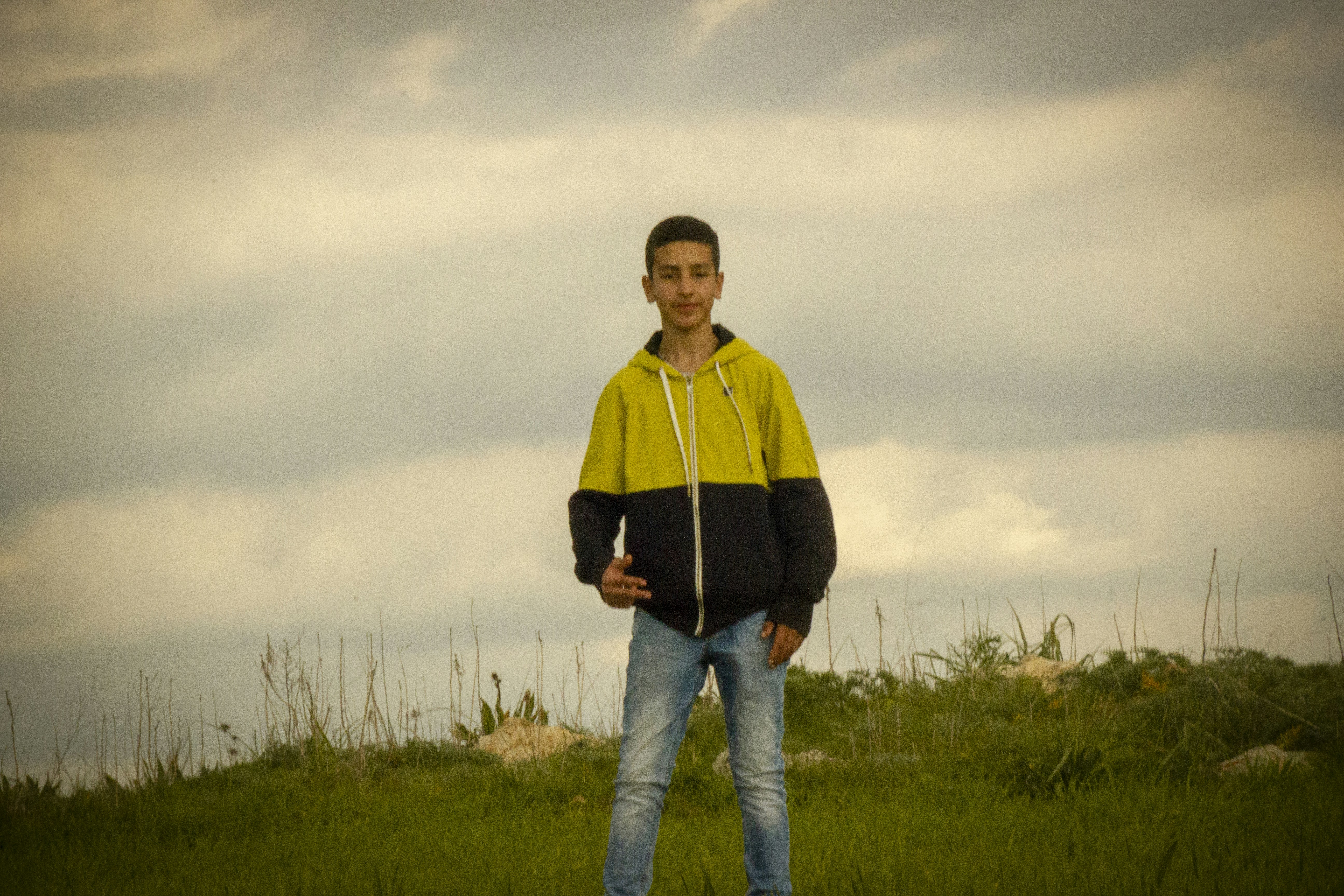 A young boy stands confidently on a grassy hill under a cloudy sky, showcasing a blend of nature and youthful energy.
