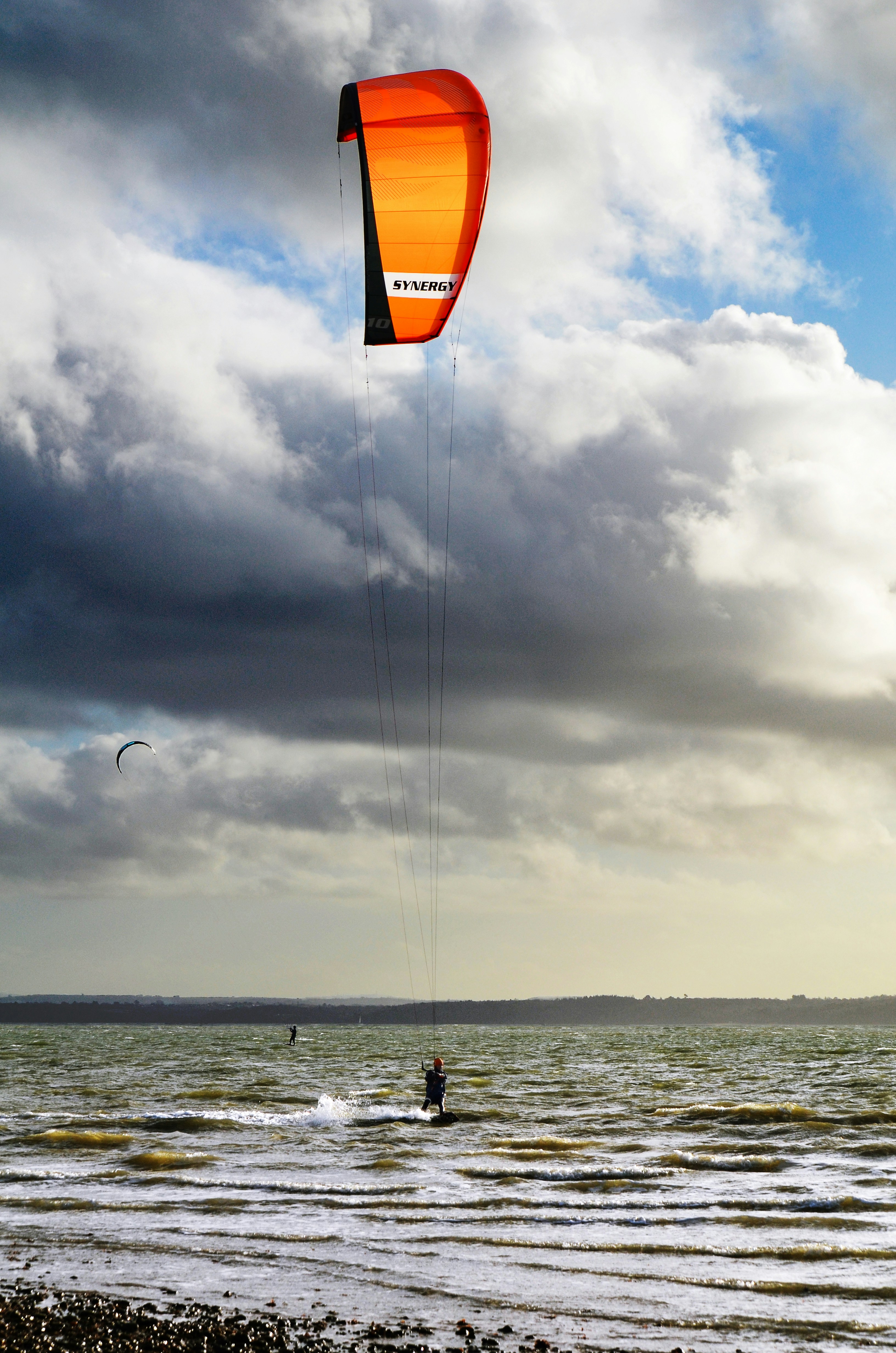 Kiteboarder gliding across choppy waters beneath a dramatic sky with a vibrant orange kite. Waves crash gently as the sun breaks through the clouds.