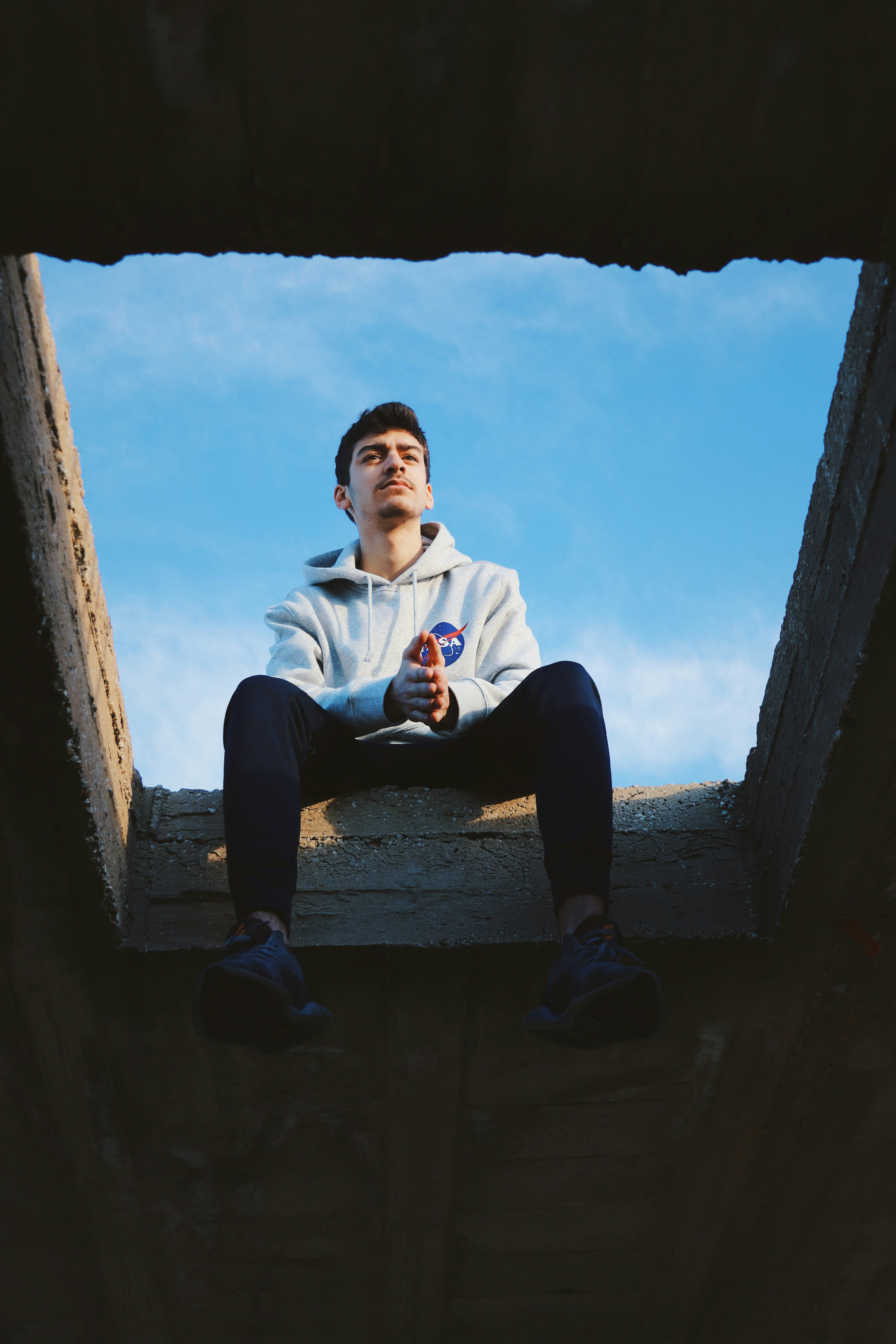 Young man seated in a concrete structure, gazing thoughtfully at the sky through an opening. The scene captures a blend of urban decay and introspective calm.