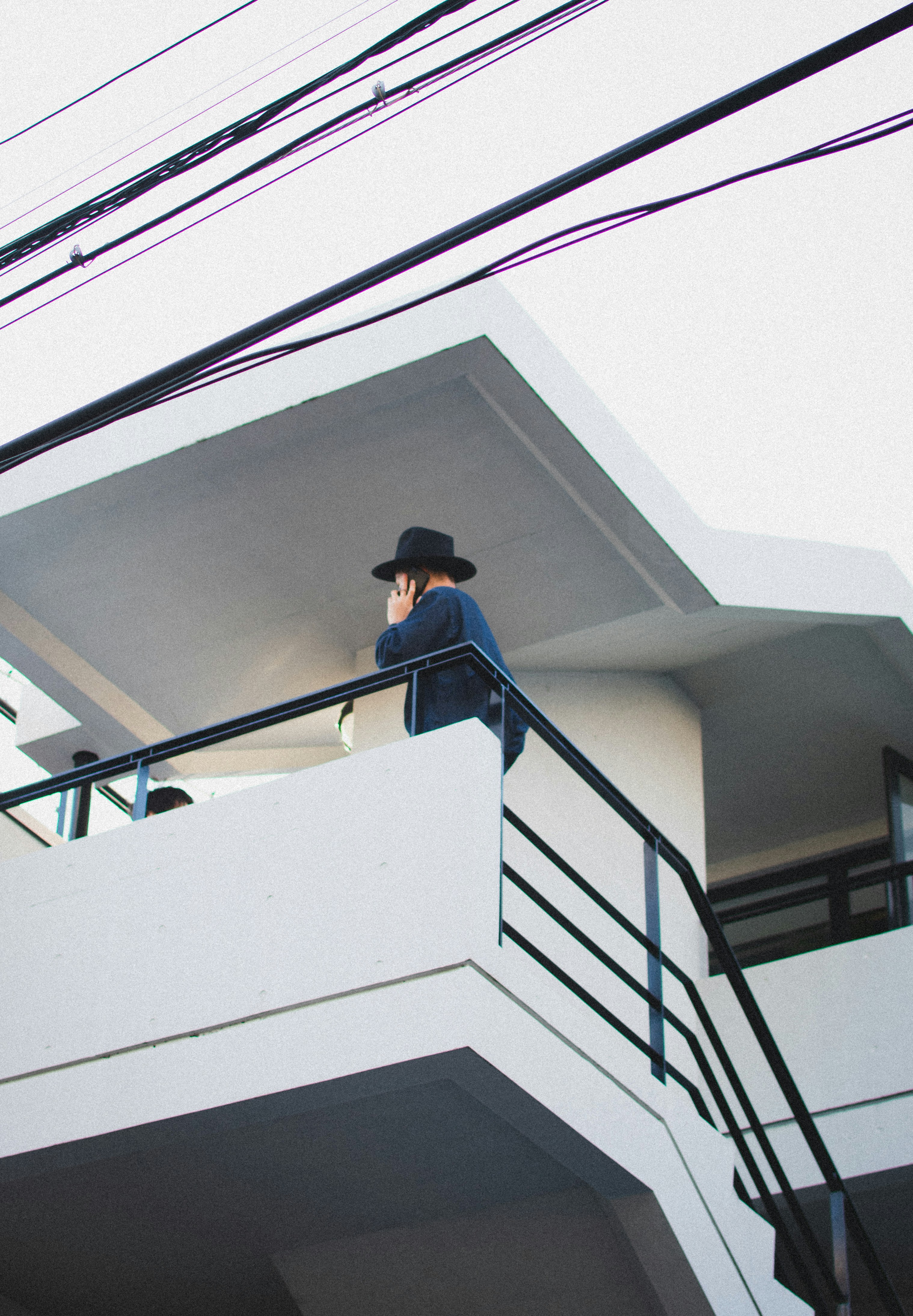 Person in a black hat stands on a balcony, engaged in a phone conversation amidst a backdrop of minimalist architecture.