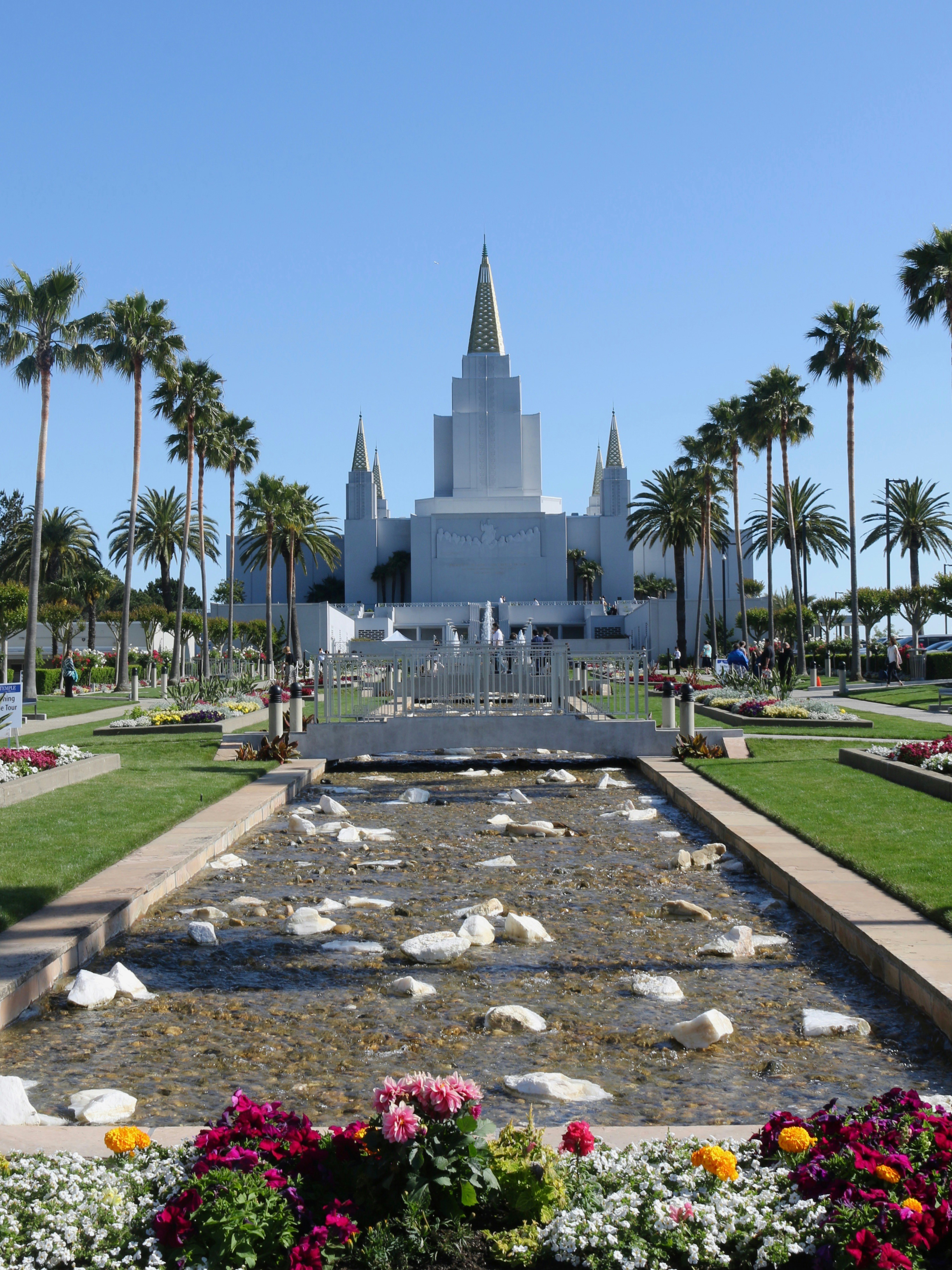 White concrete temple with gold spires framed by palm trees and a vibrant garden, under a clear blue sky.
