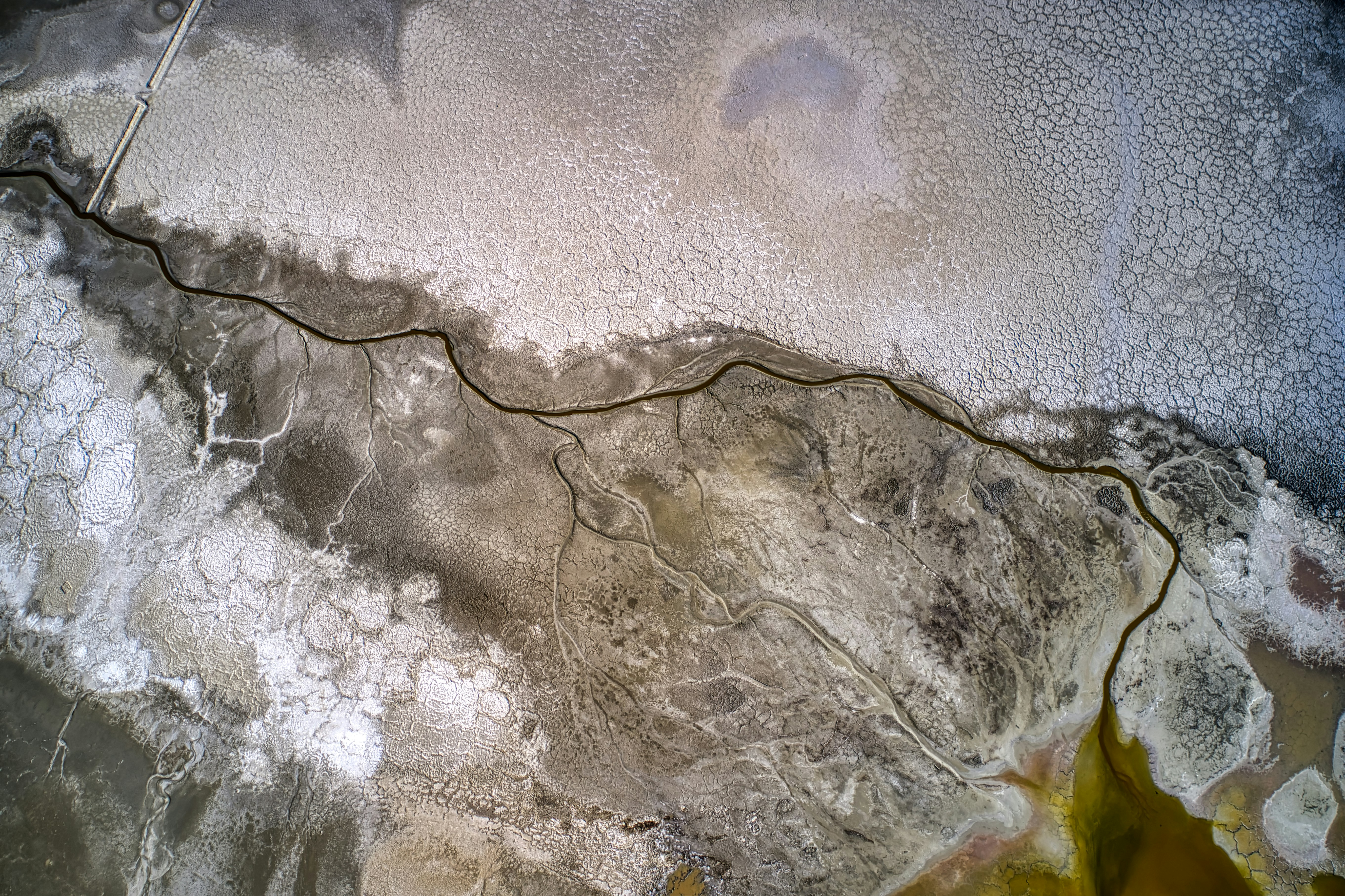 Aerial view of a winding waterway traversing a desolate salt flat, showcasing intricate patterns and textures in the earth. The stark contrast of colors adds depth to the barren terrain.