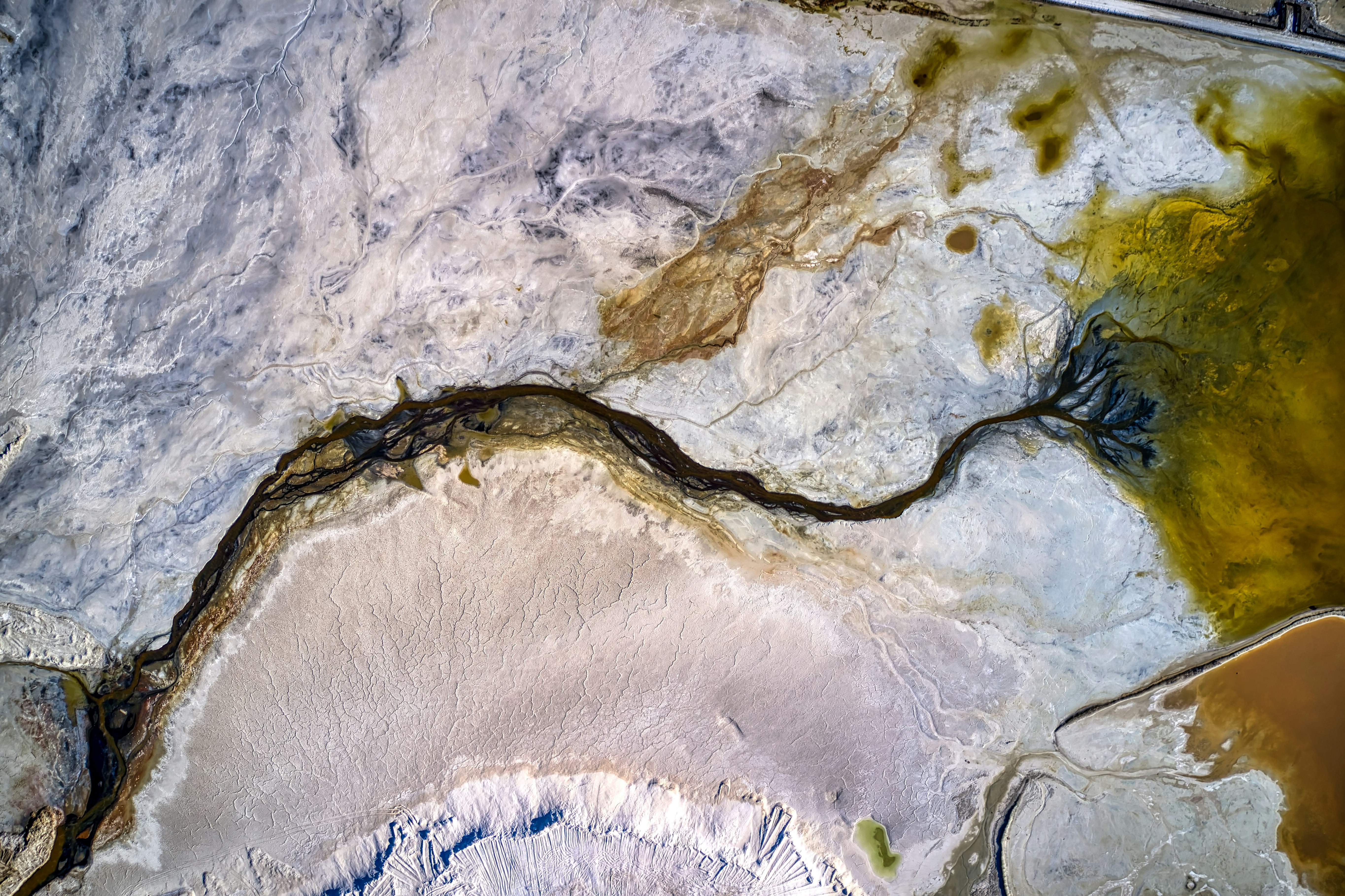 Abstract aerial view of a winding dark channel through salt flats, showcasing intricate patterns and contrasting colors of earth and water.
