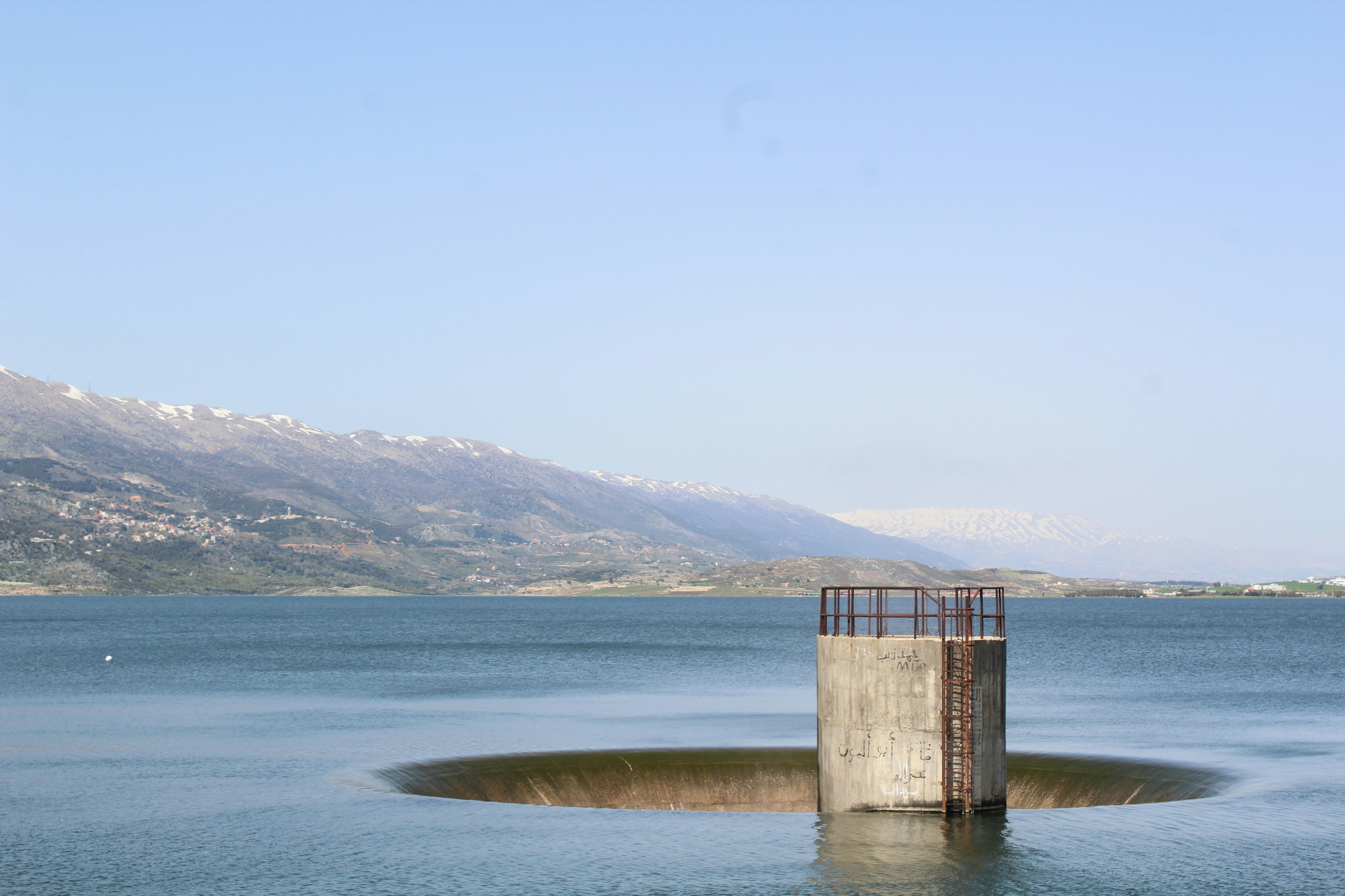 Concrete structure emerging from tranquil water, surrounded by distant mountains under a clear sky.