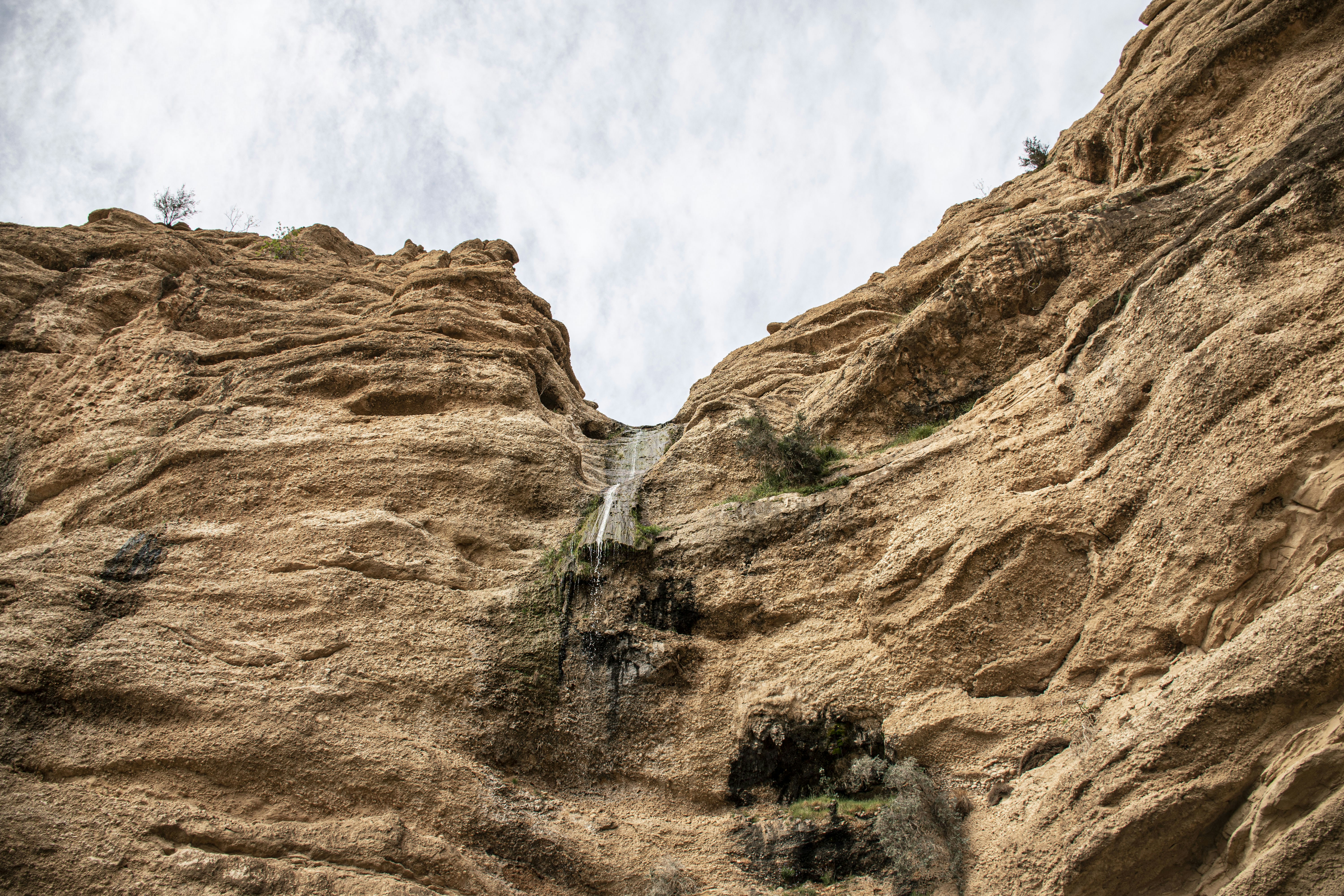 A waterfall cascading down rugged sandstone cliffs under a cloudy sky, showcasing the intricate textures of the rock face.