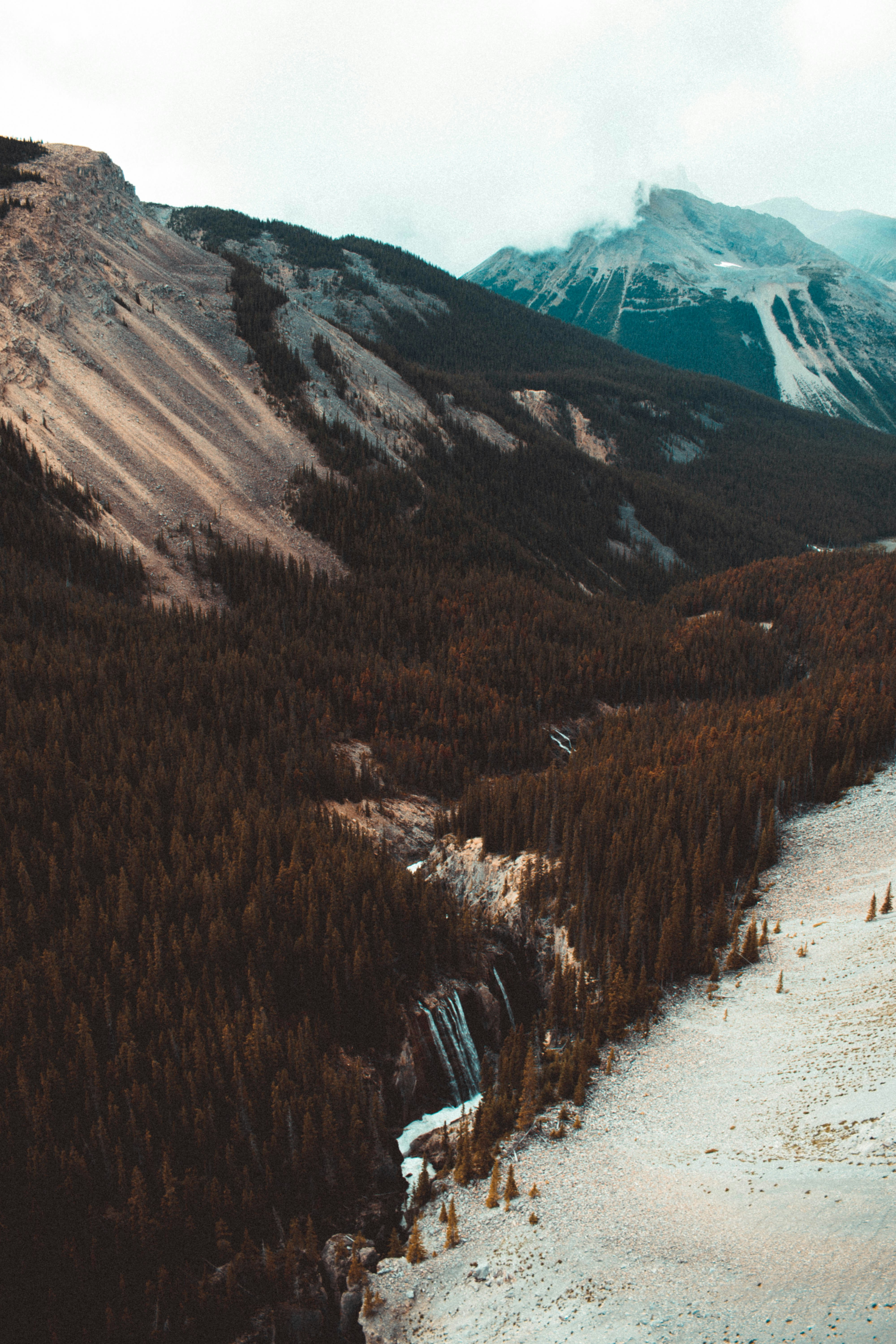 Aerial view of a cascading waterfall nestled among dense evergreen forests and rugged mountain terrain, showcasing the serene beauty of nature.