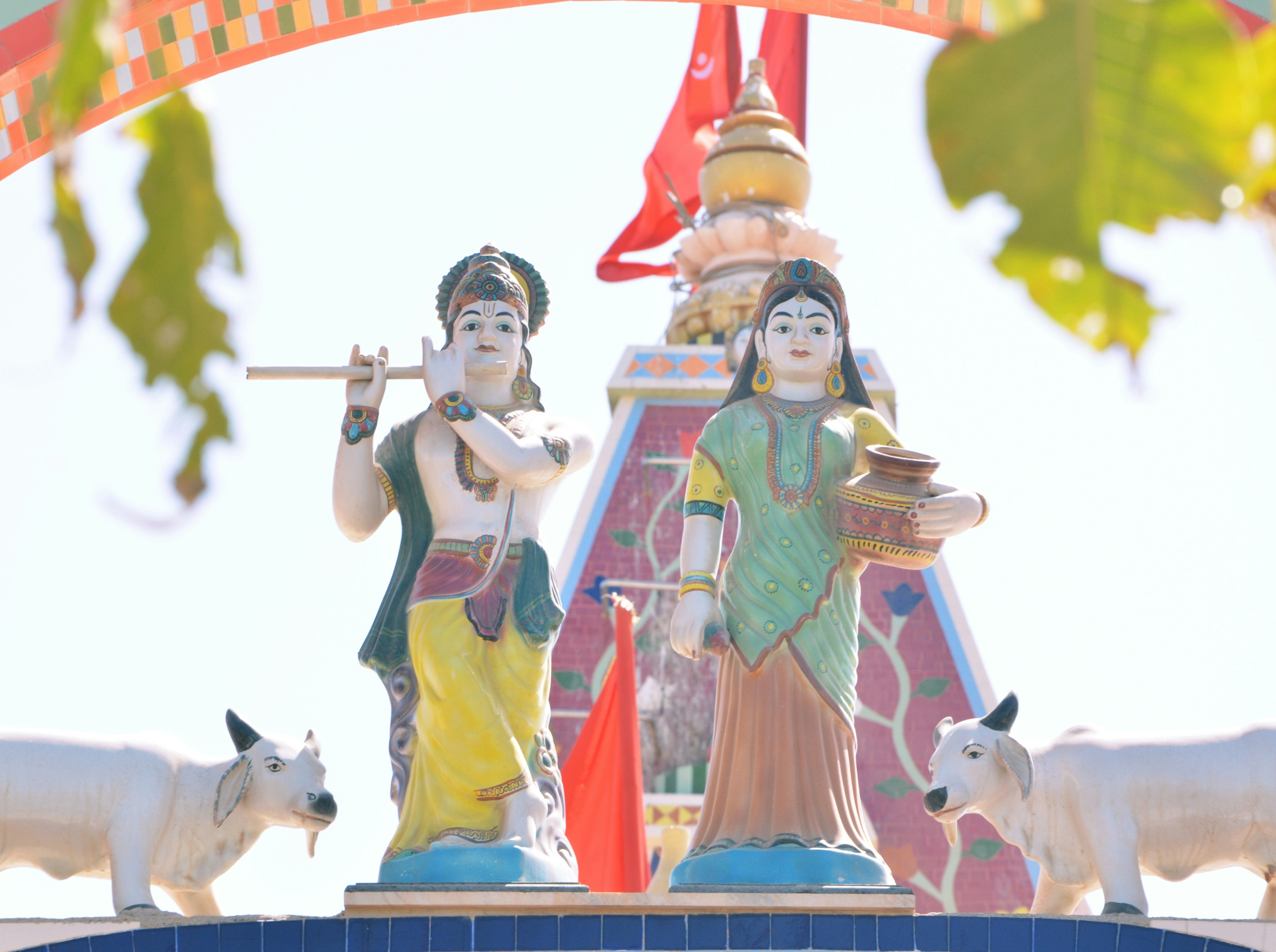 Colorful statues of two deities with cows beneath a decorative archway, surrounded by leaves.
