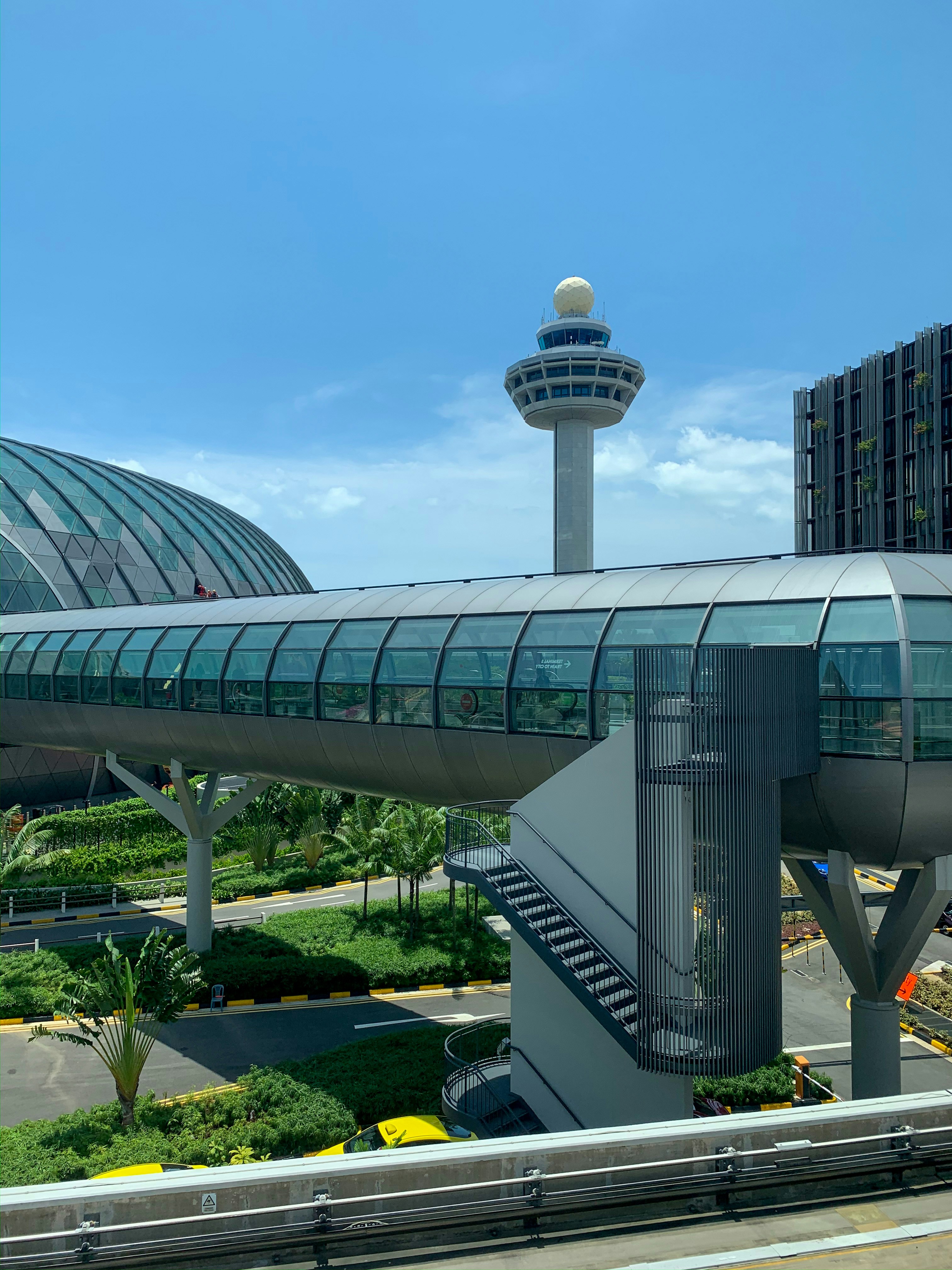 A sleek, glass-enclosed walkway connects two contemporary structures near an airport, with a control tower visible in the background. Lush greenery frames the scene.