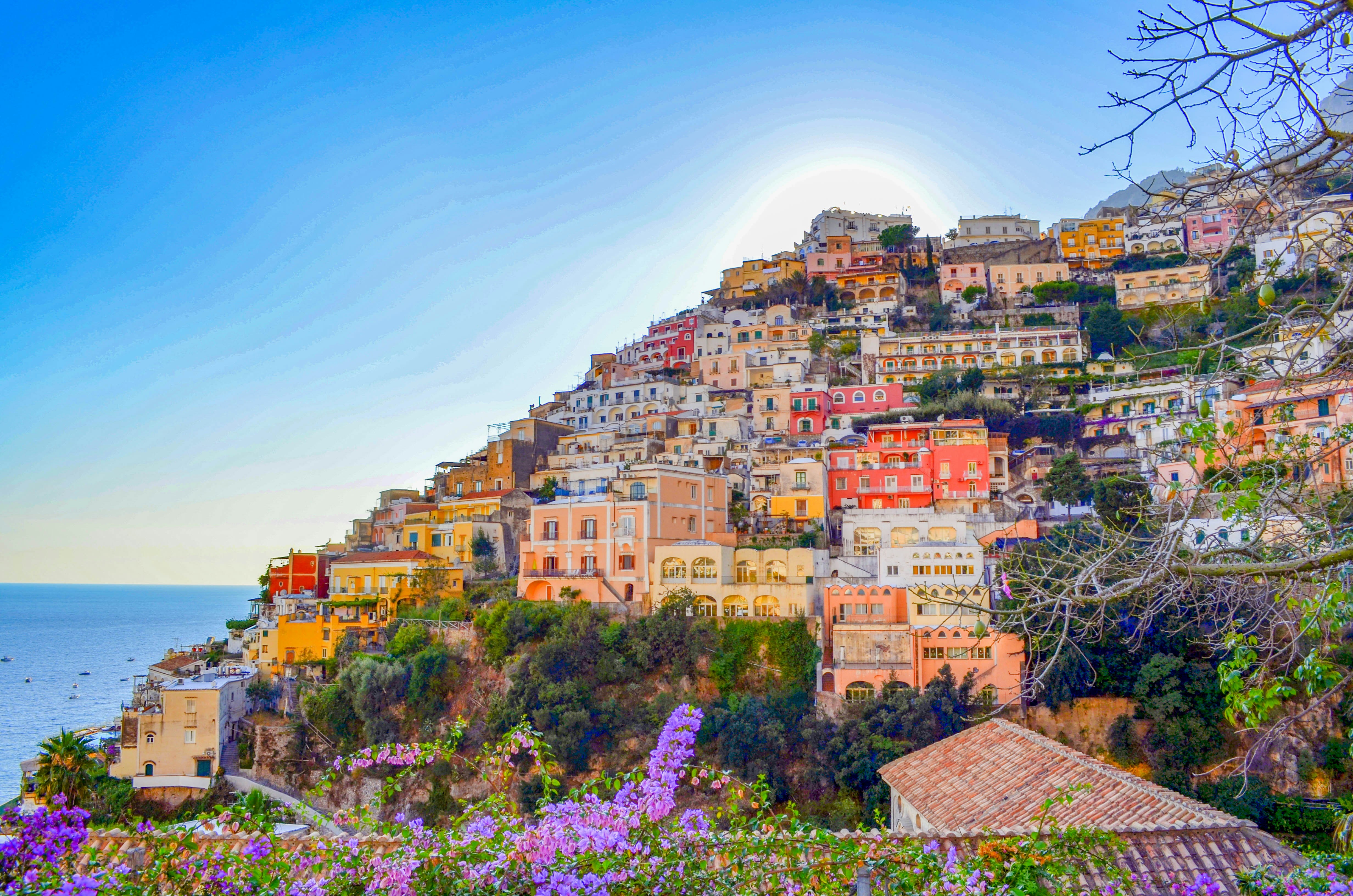 Colorful buildings cascade down a hillside overlooking the serene Mediterranean Sea, adorned with blooming flowers in the foreground.