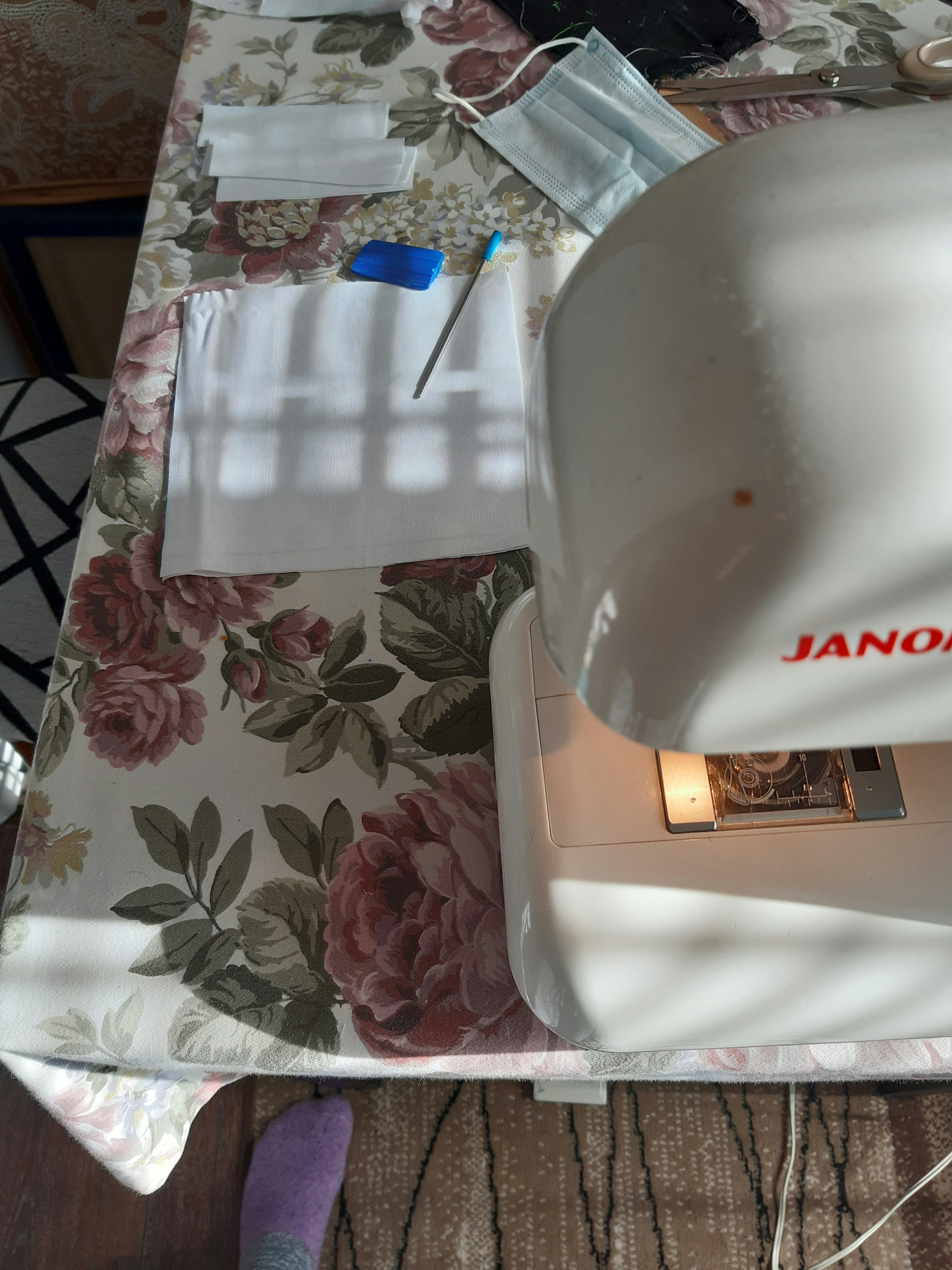 Sewing machine positioned on a floral tablecloth, surrounded by tools and materials for crafting. Sunlight casts shadows, enhancing the workspace atmosphere.