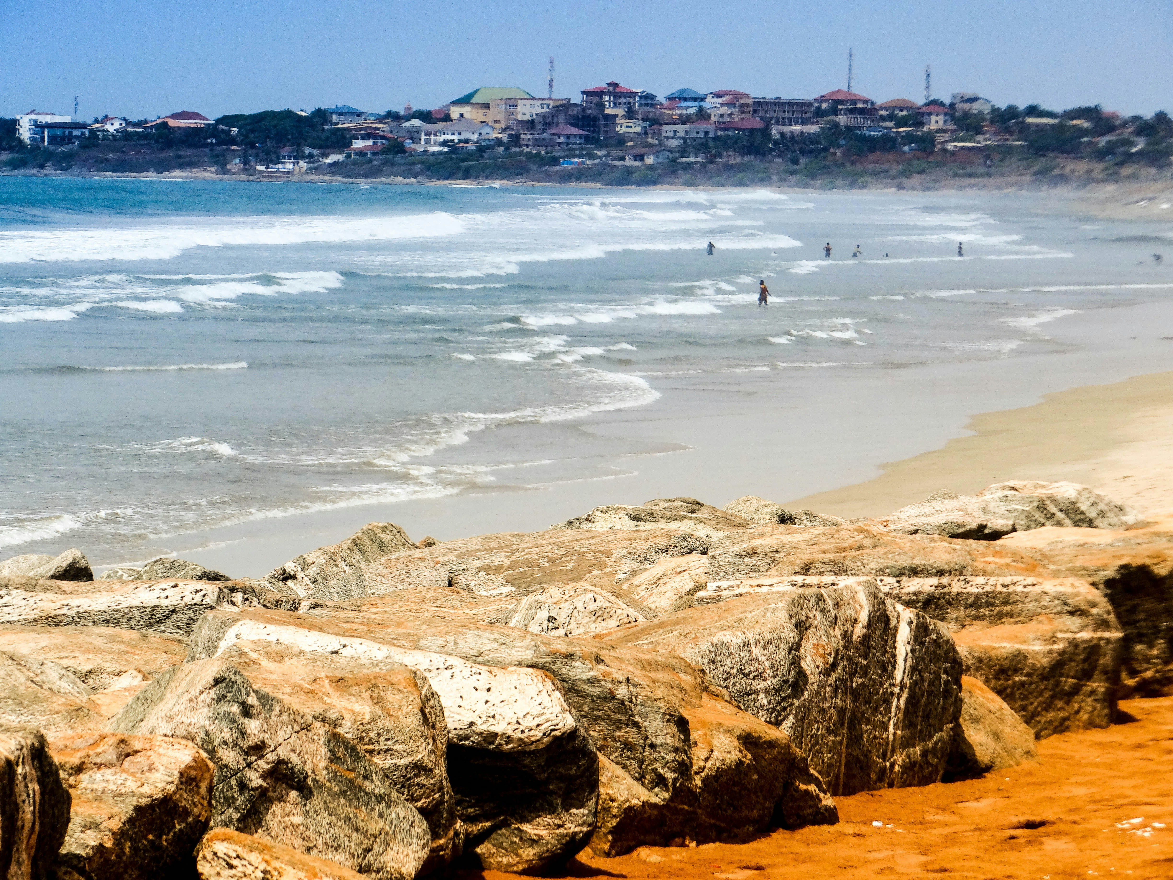 Rugged coastline with large rocks in the foreground and waves lapping against the sandy shore under a clear sky.