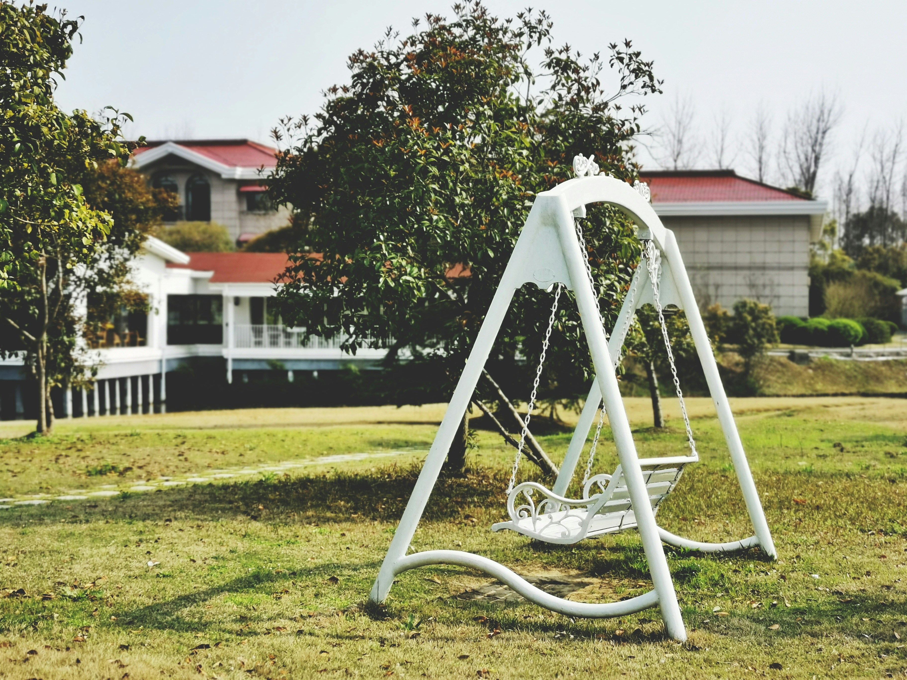 A vintage white swing set nestled in a lush green garden, framed by a charming house in the background. The scene evokes a sense of tranquility and nostalgia.