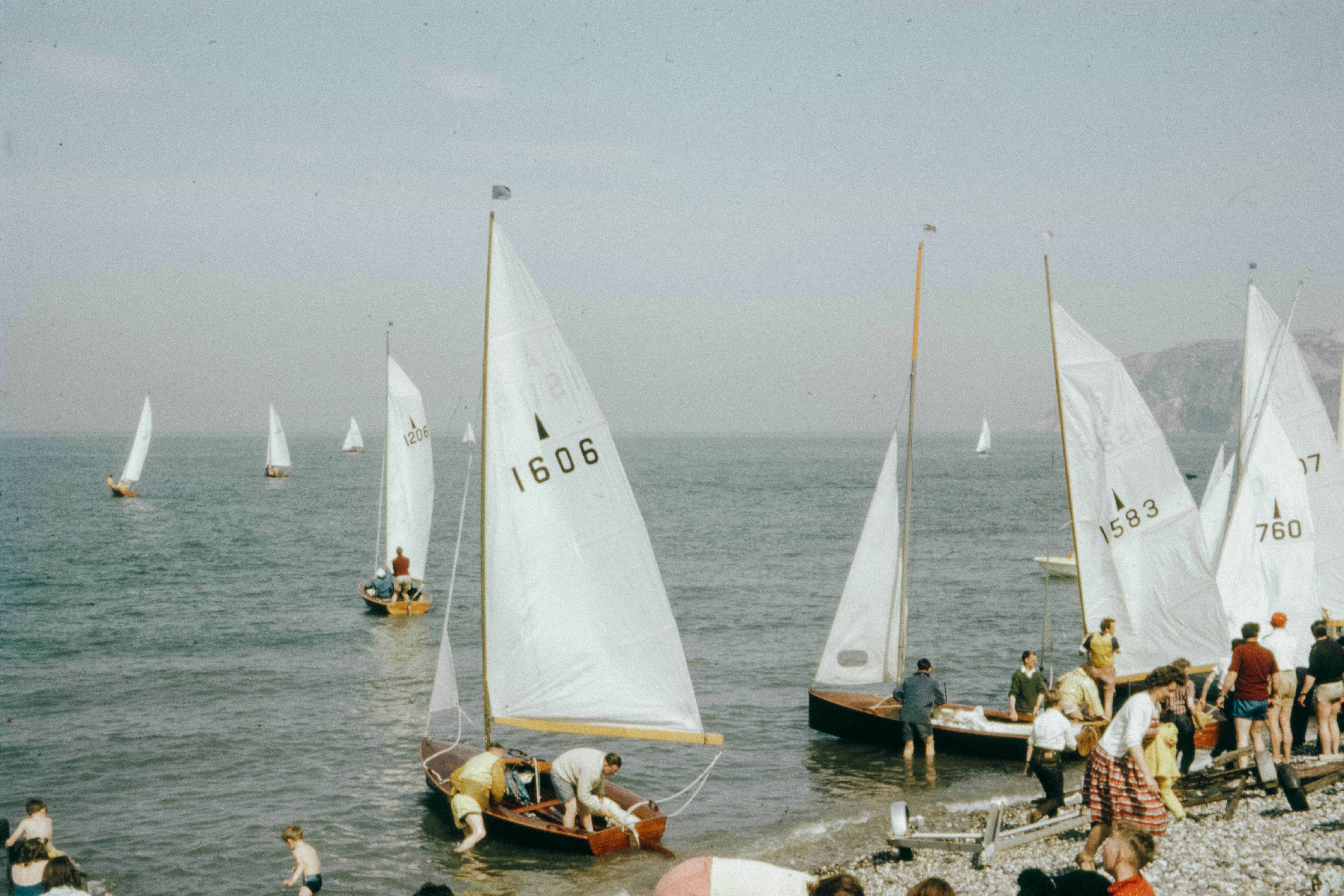 Bustling beach scene with several small sailboats near the shore and groups of sunbathers along the water's edge.