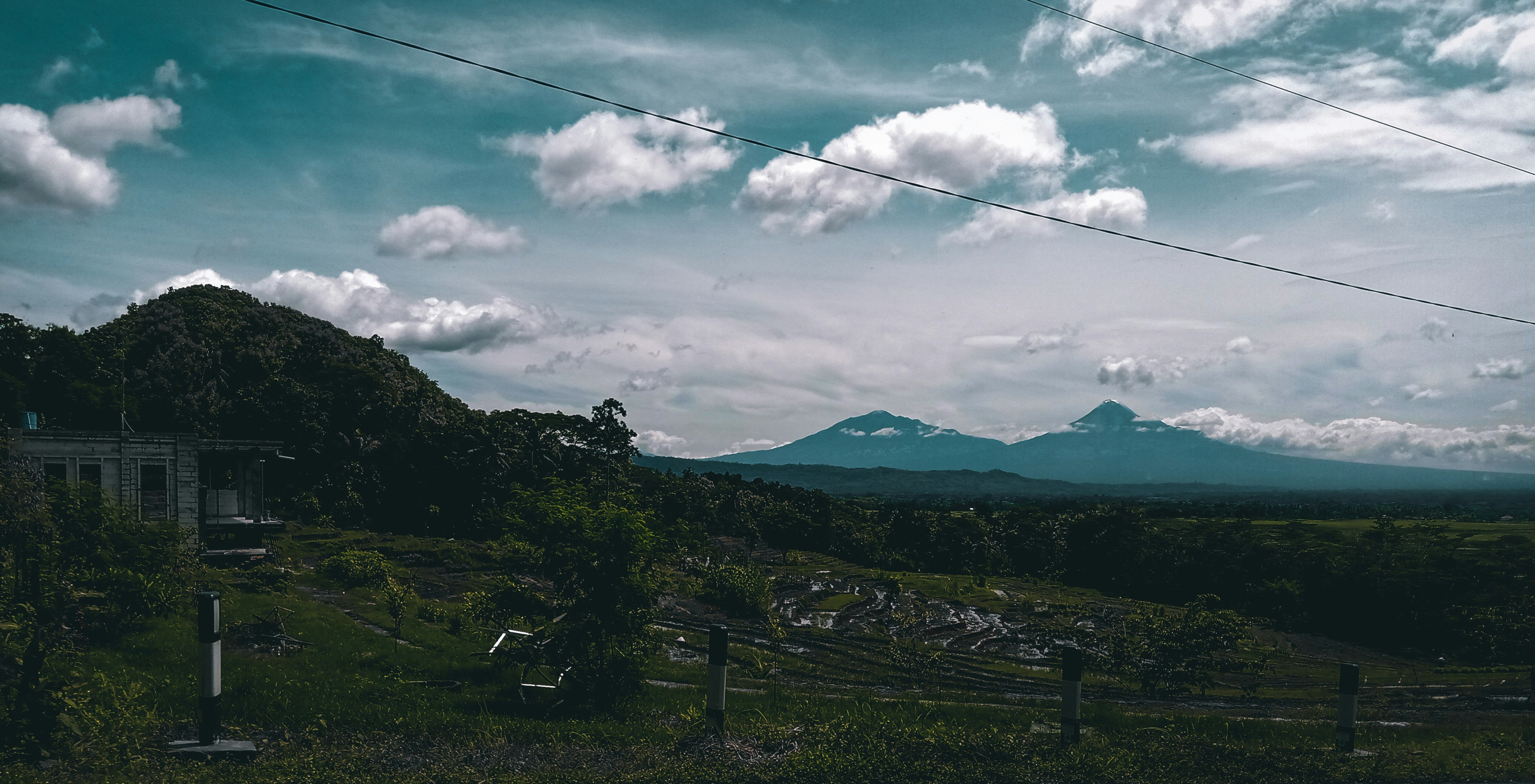 Lush green hills under a moody sky, with distant mountains looming on the horizon. A rustic structure adds depth to the tranquil scene.