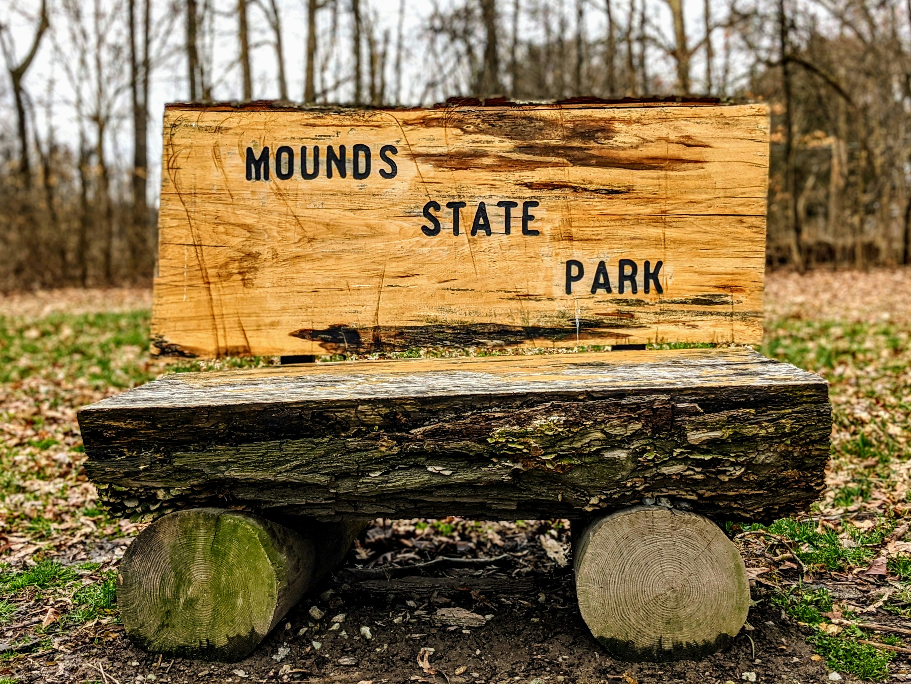 Wooden bench with 'Mounds State Park' engraved, set against a backdrop of bare trees.
