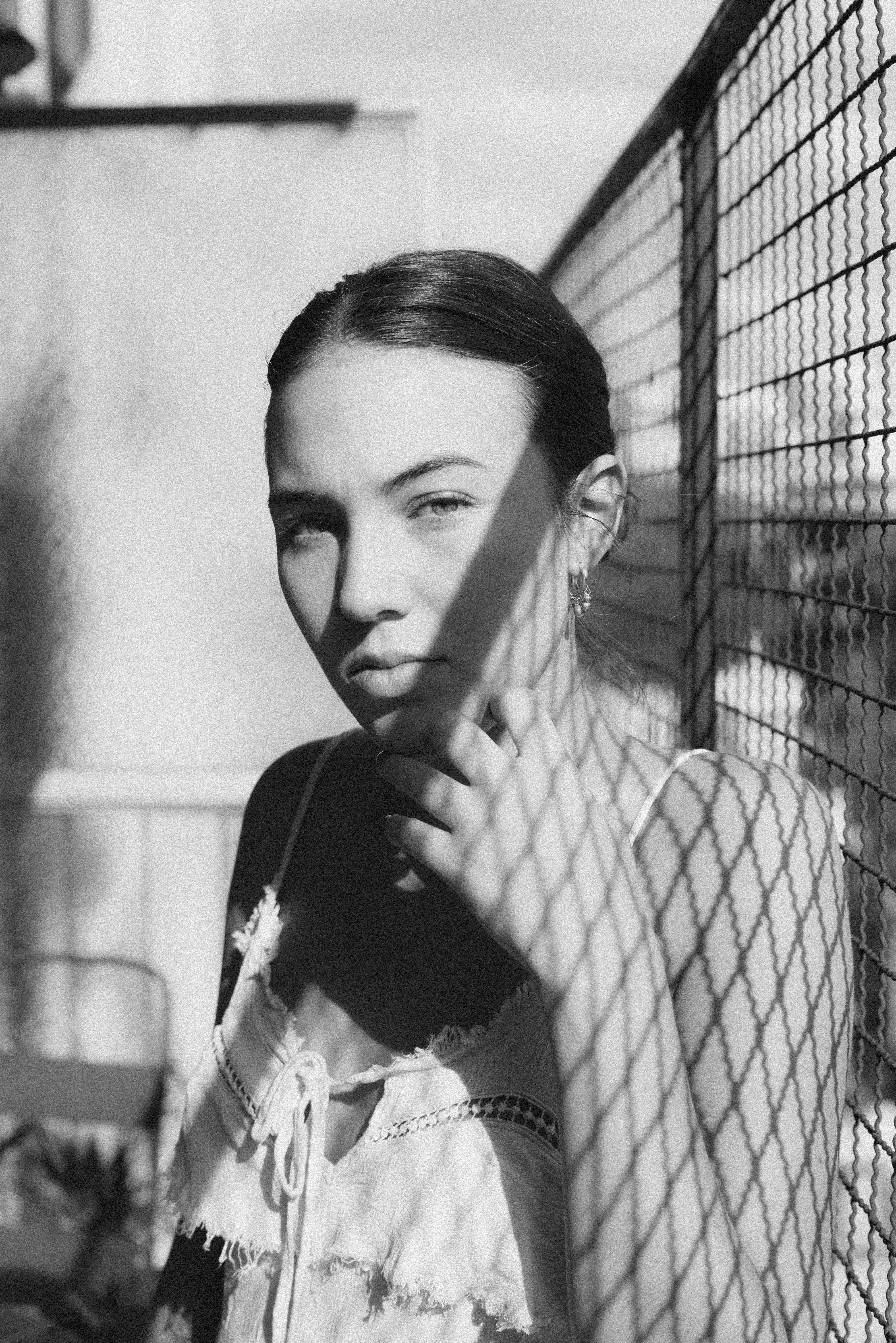 A young woman gazes thoughtfully, her face partially illuminated by sunlight filtered through a patterned fence. The interplay of light and shadow creates a striking contrast.