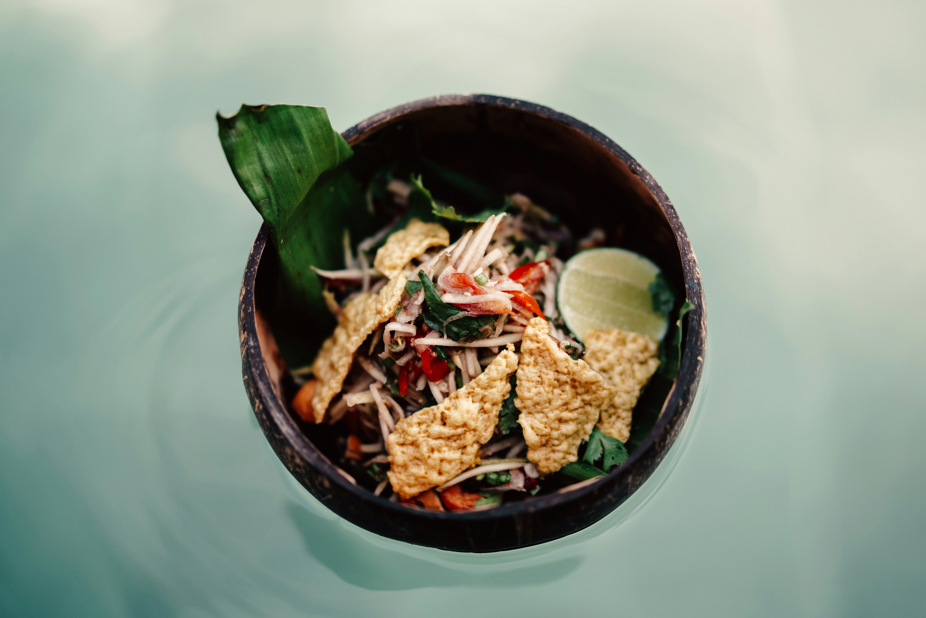 Vibrant salad with fresh herbs and crispy toppings served in a coconut bowl, floating on tranquil water.